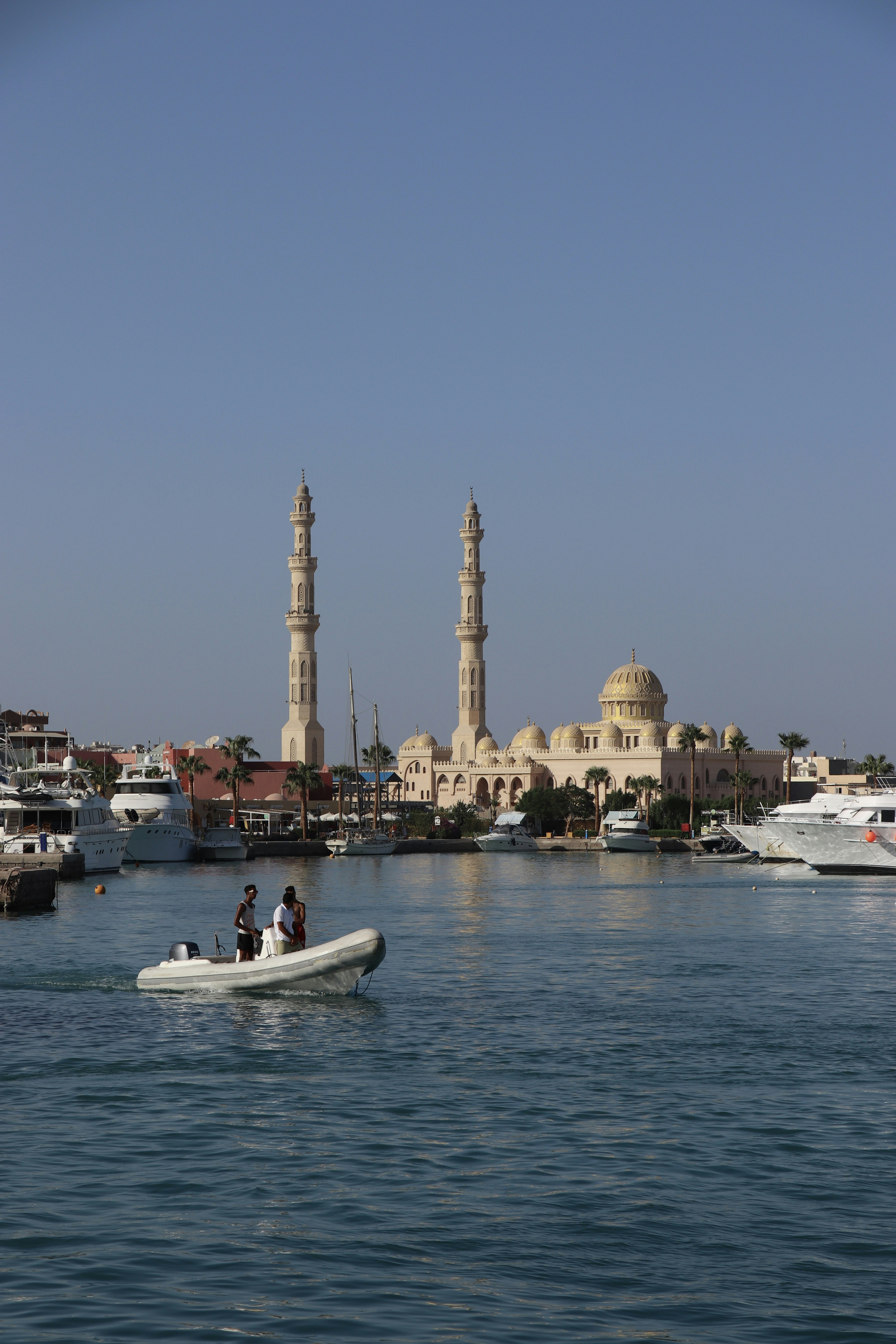 A lively scene in the Hurghada marina, Egypt, where a small motorboat glides across the calm waters in front of the grand Abdulmoneim Riadh Mosque. The golden domes and towering minarets form a striking contrast with the deep blue sea and the luxury yachts docked along the promenade. A modern coastal moment captured beside timeless architecture. | A mosque stands tall over a vibrant harbor.