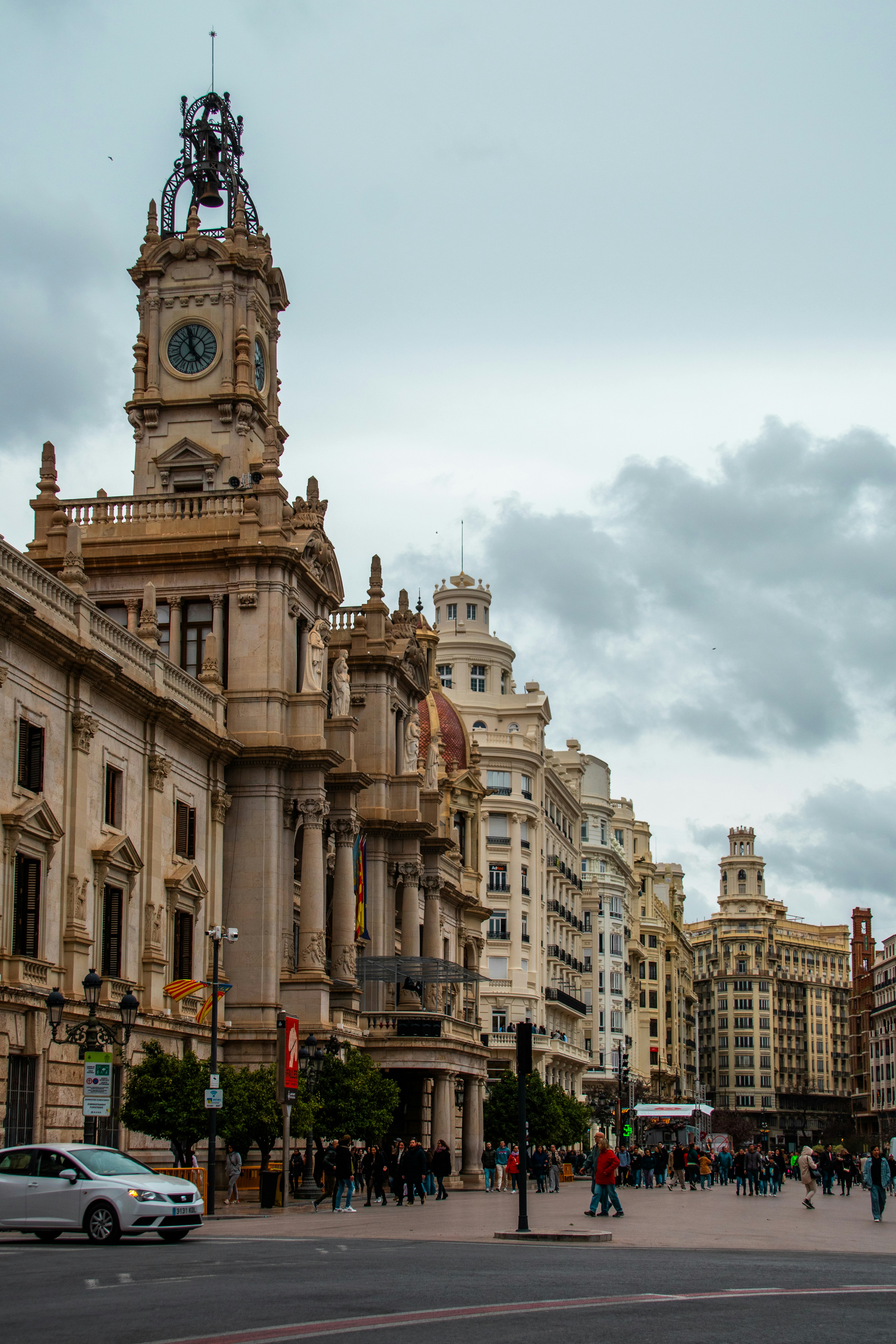 Historic buildings line a bustling city street under a cloudy sky, capturing the vibrant life of urban architecture.