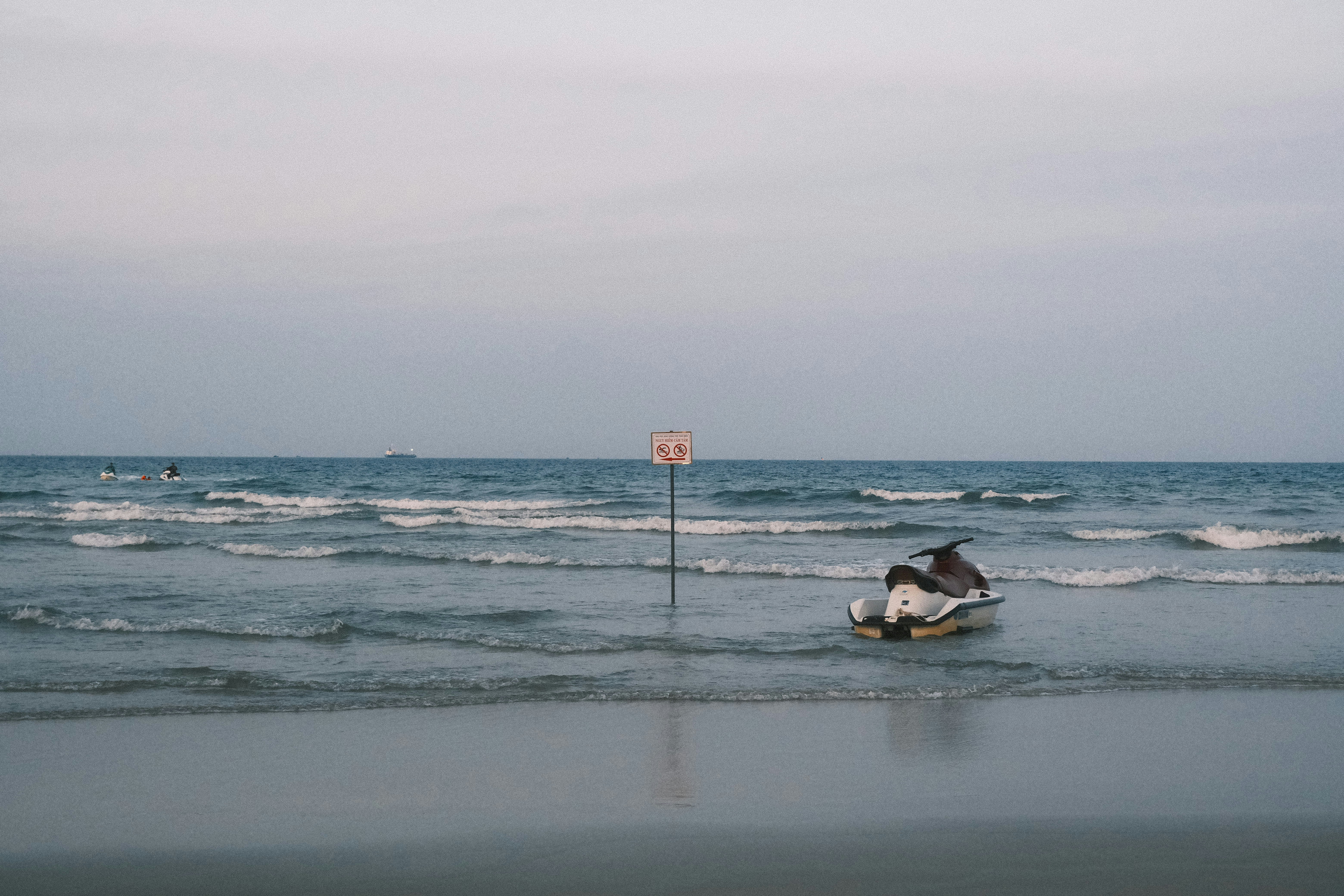 A jet ski rests near a warning sign on a tranquil beach, with gentle waves lapping at the shore. The horizon fades into a soft, pastel sky.