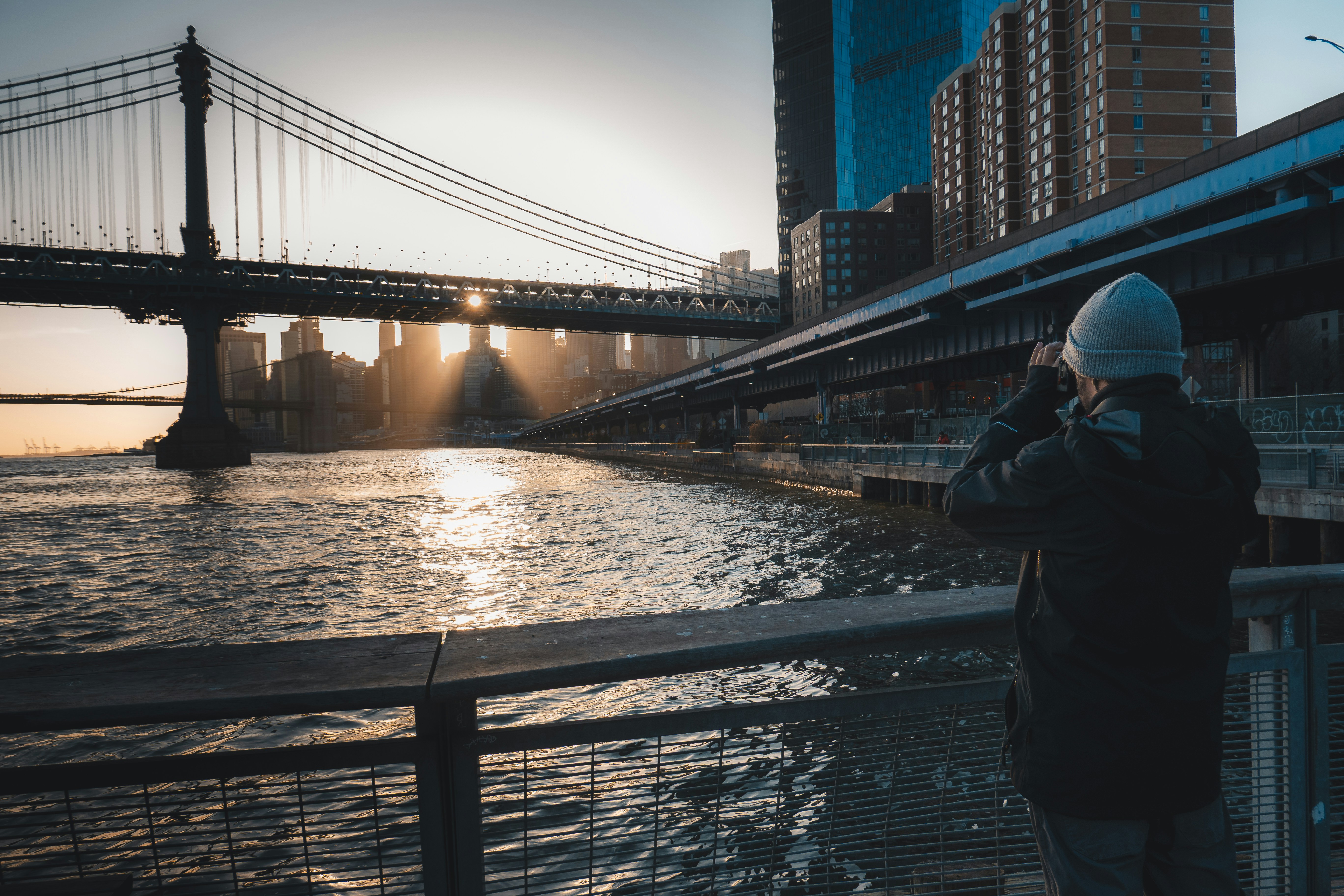 A photographer capturing the golden hour near a bridge, with sunlight reflecting off the water and urban buildings in the background.