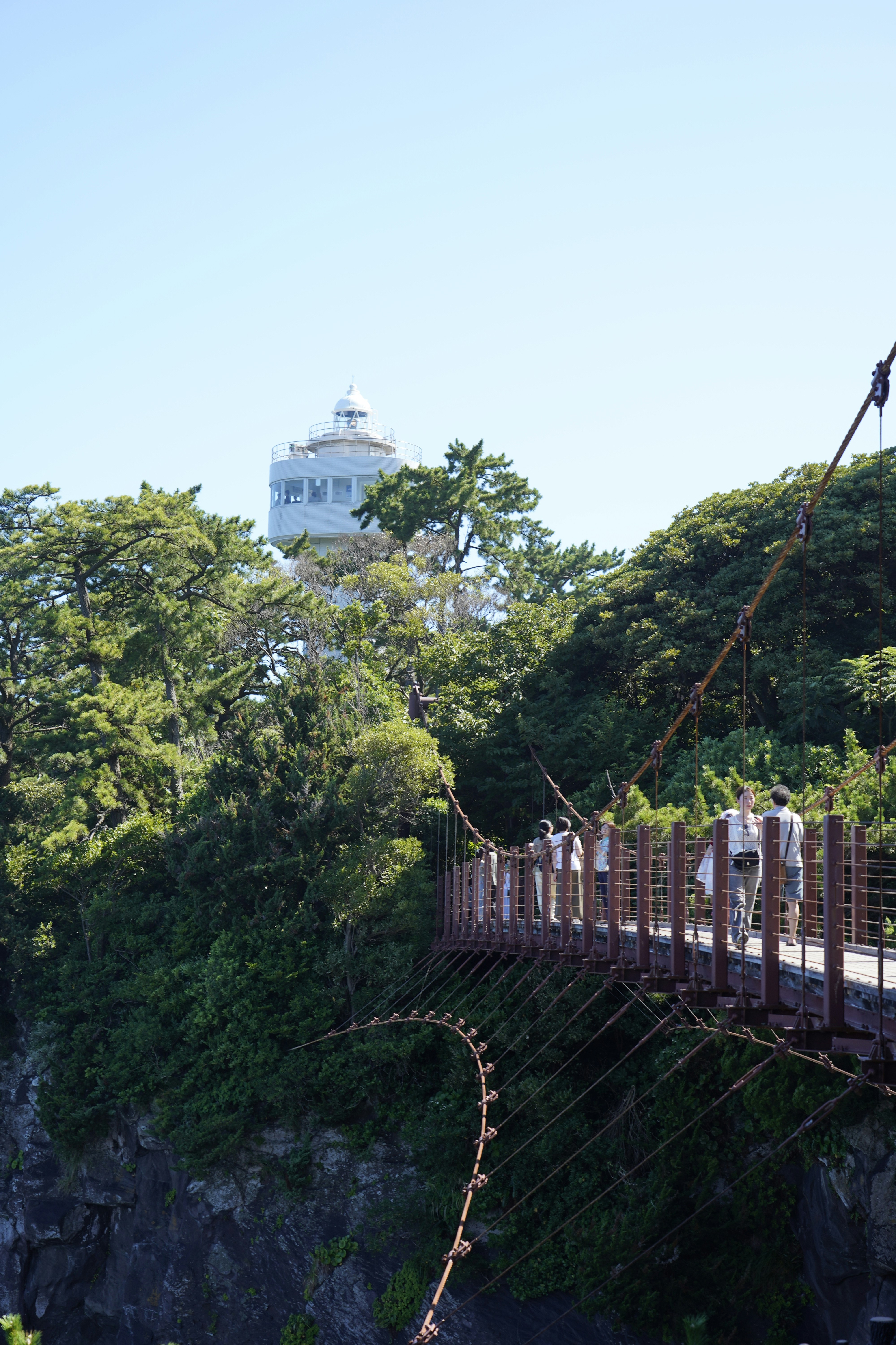Visitors traverse a suspension bridge surrounded by lush greenery, with a lighthouse peeking through the trees in the background.