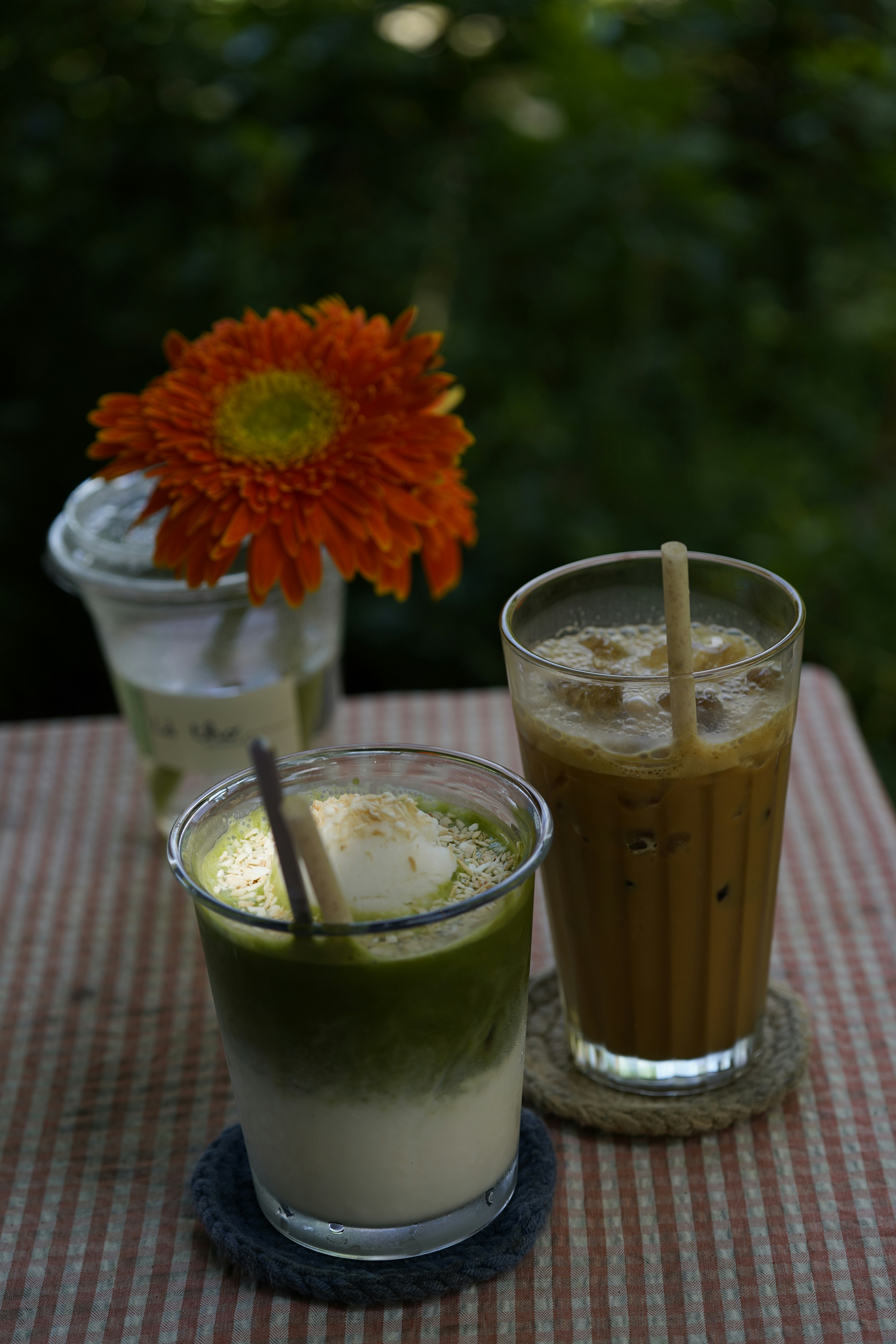 Two glasses of iced beverages, one with a matcha layer and the other a classic iced coffee, accompanied by a vibrant orange flower in a jar. A cozy setting enhances the inviting atmosphere.