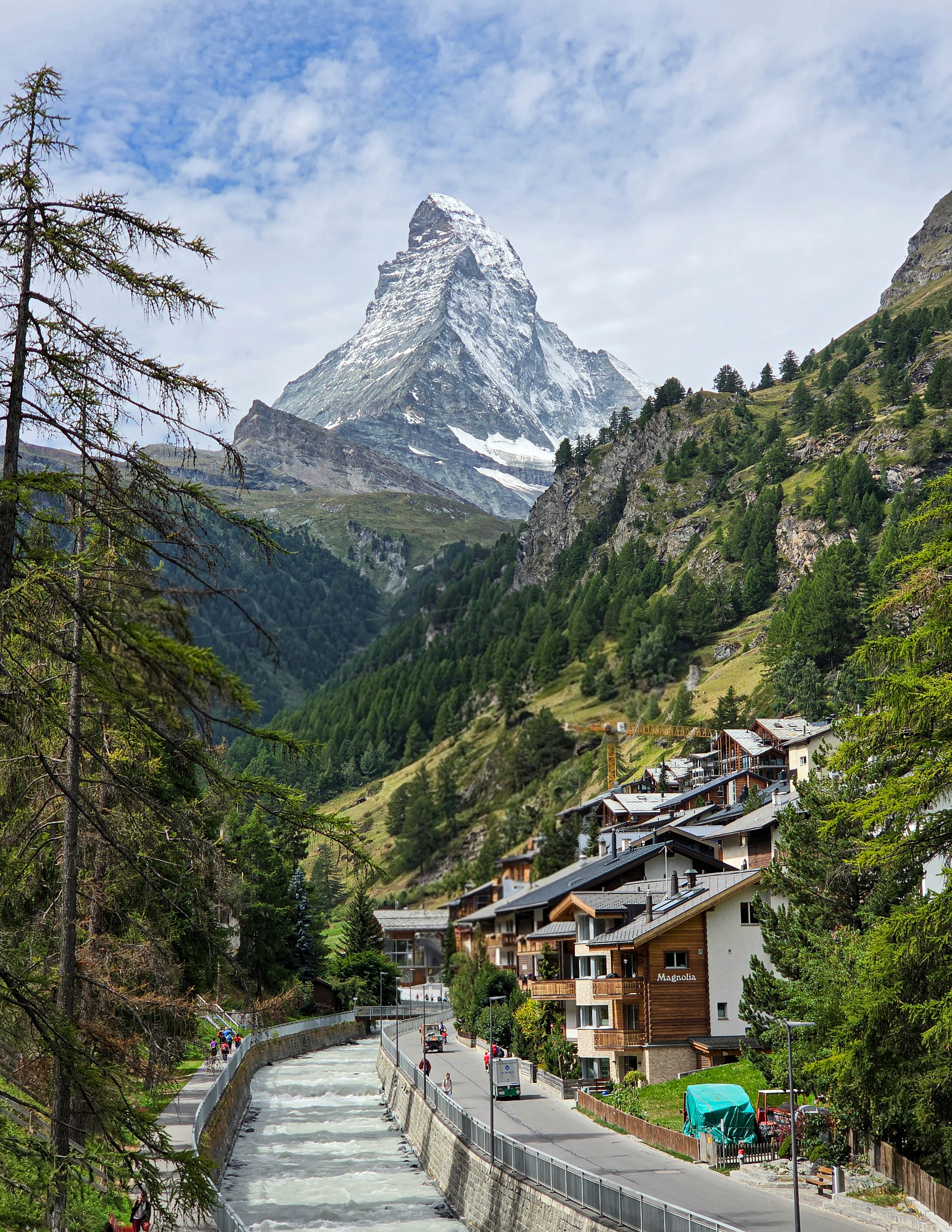 The Matterhorn towers over charming alpine houses along a winding river, framed by lush greenery and rugged mountains. A tranquil scene of nature and architecture harmoniously coexisting.