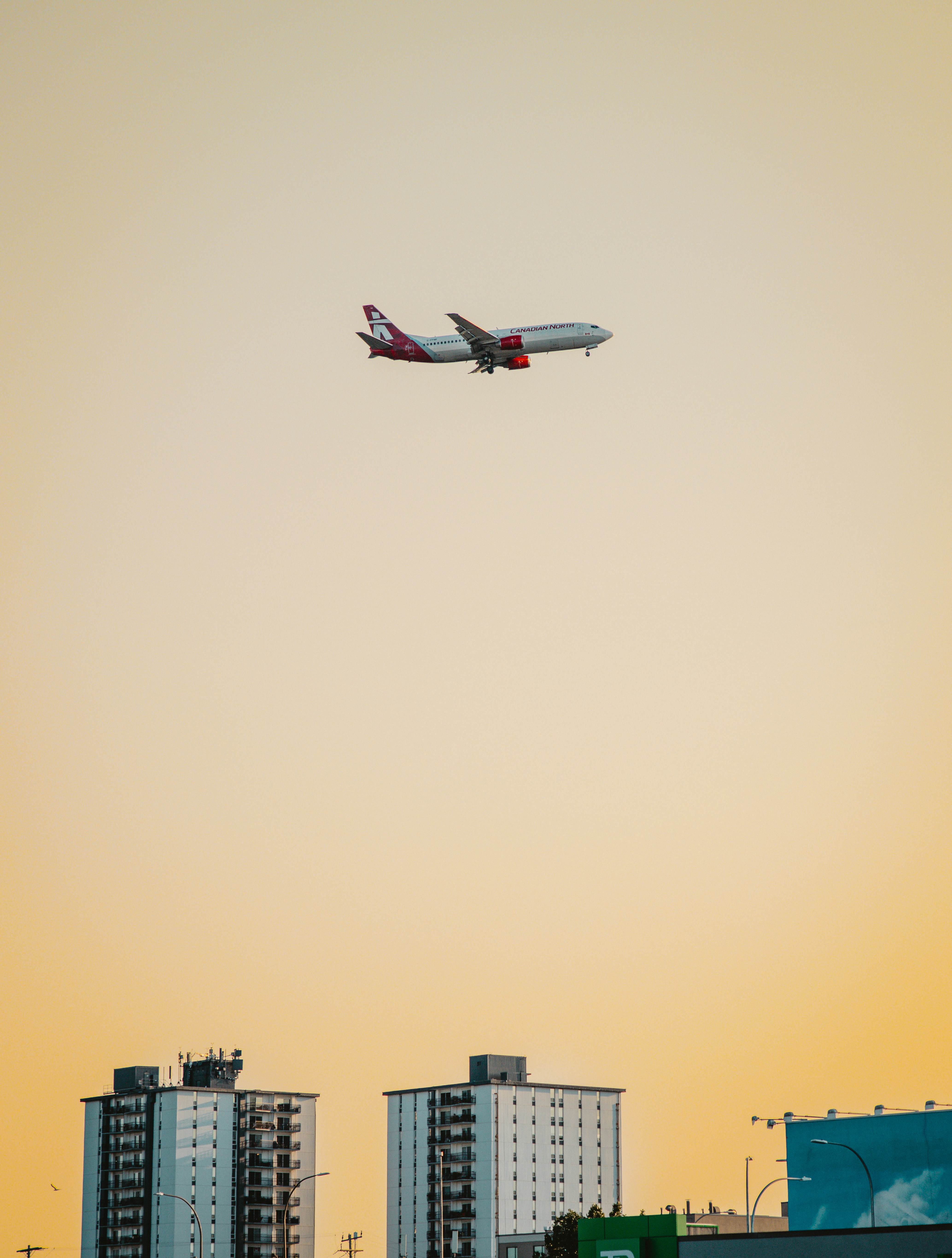 A plane flies over buildings against the sky.