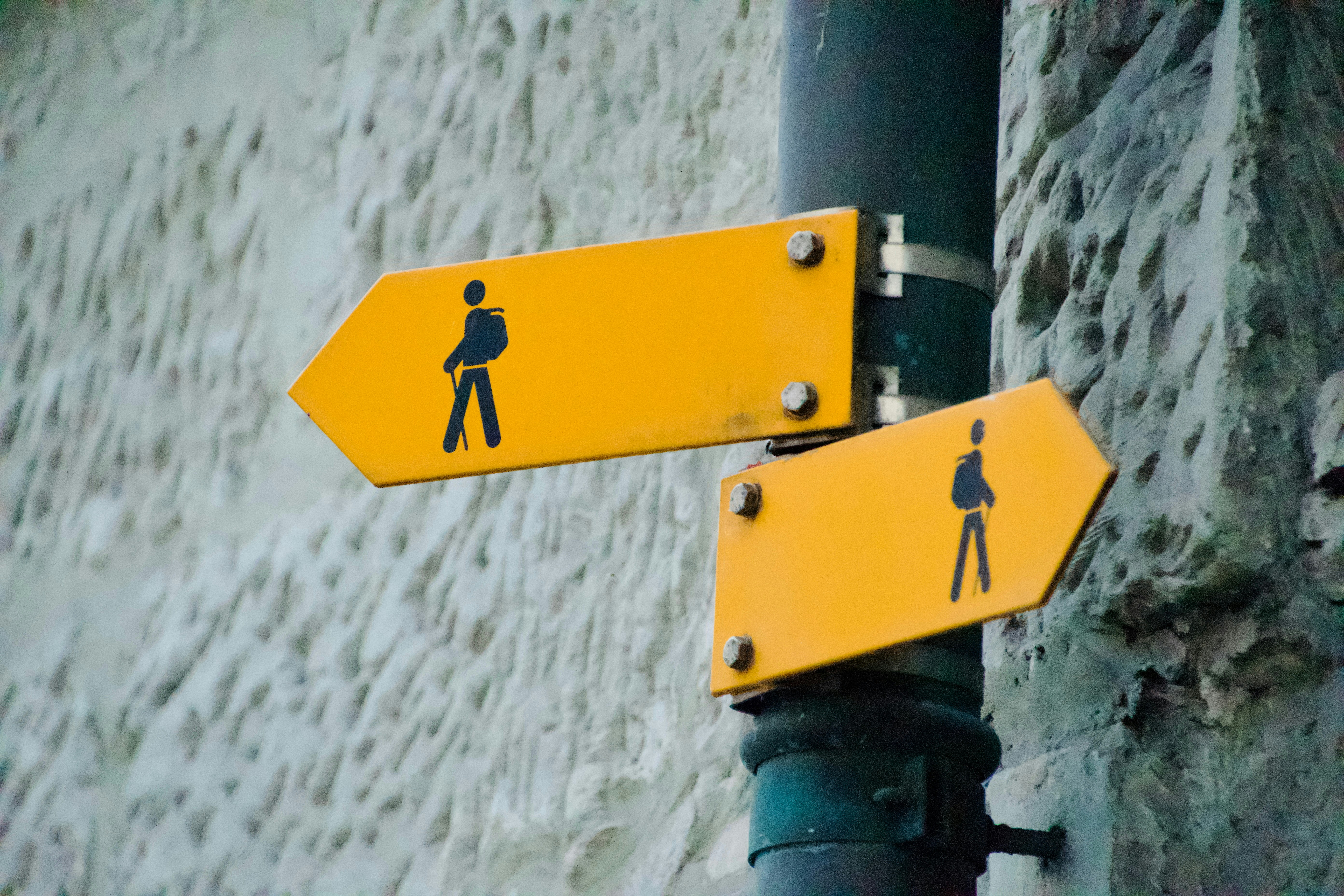 Two yellow directional signs indicating hiking paths, featuring symbols of hikers. The signs are mounted on a textured stone wall.