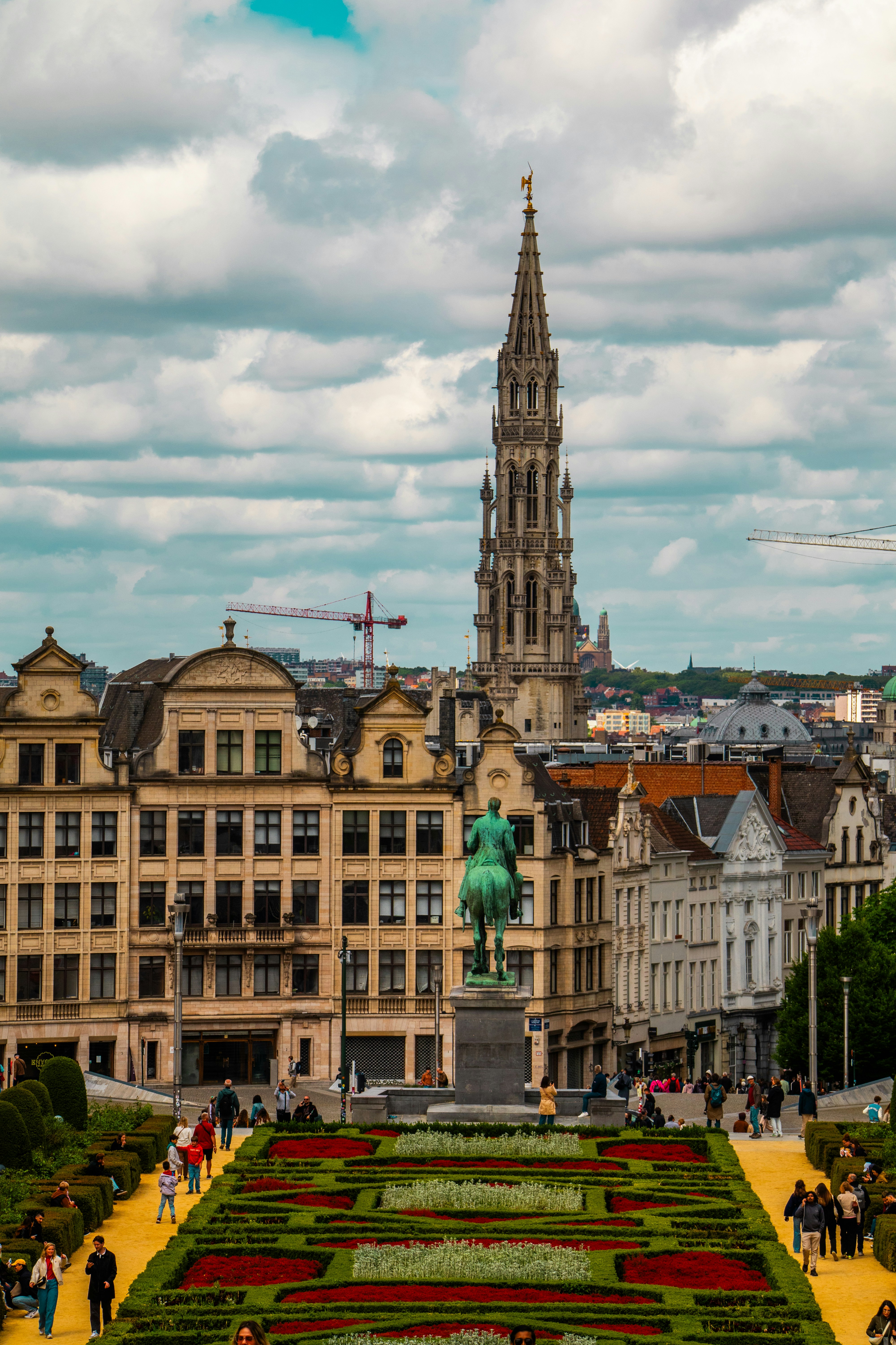 A vibrant cityscape featuring a historic statue in a meticulously designed garden, with the iconic tower of Brussels in the background under a dynamic sky.