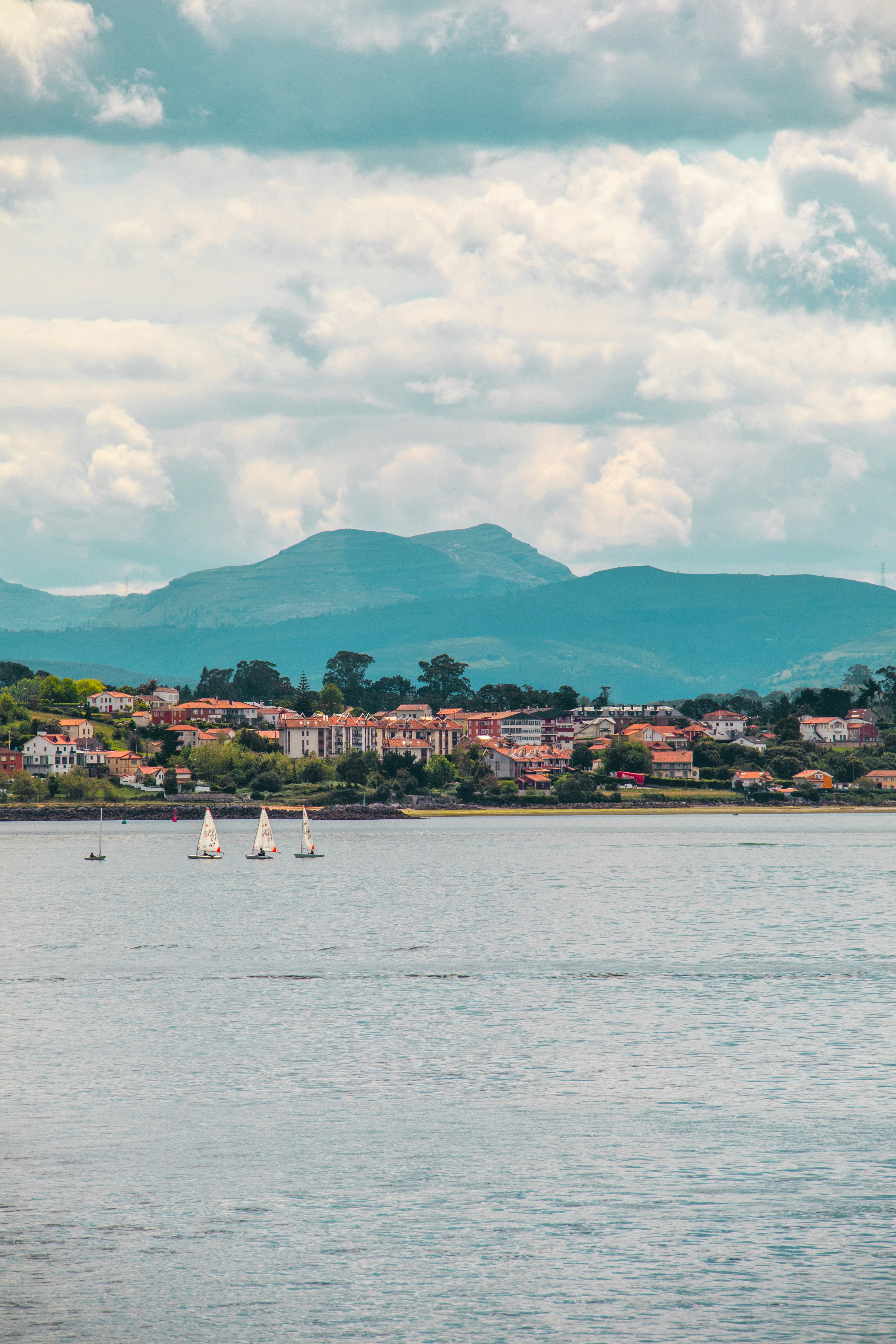 Sailboats glide across a calm bay, with a picturesque town nestled against a backdrop of rolling hills and dramatic clouds.