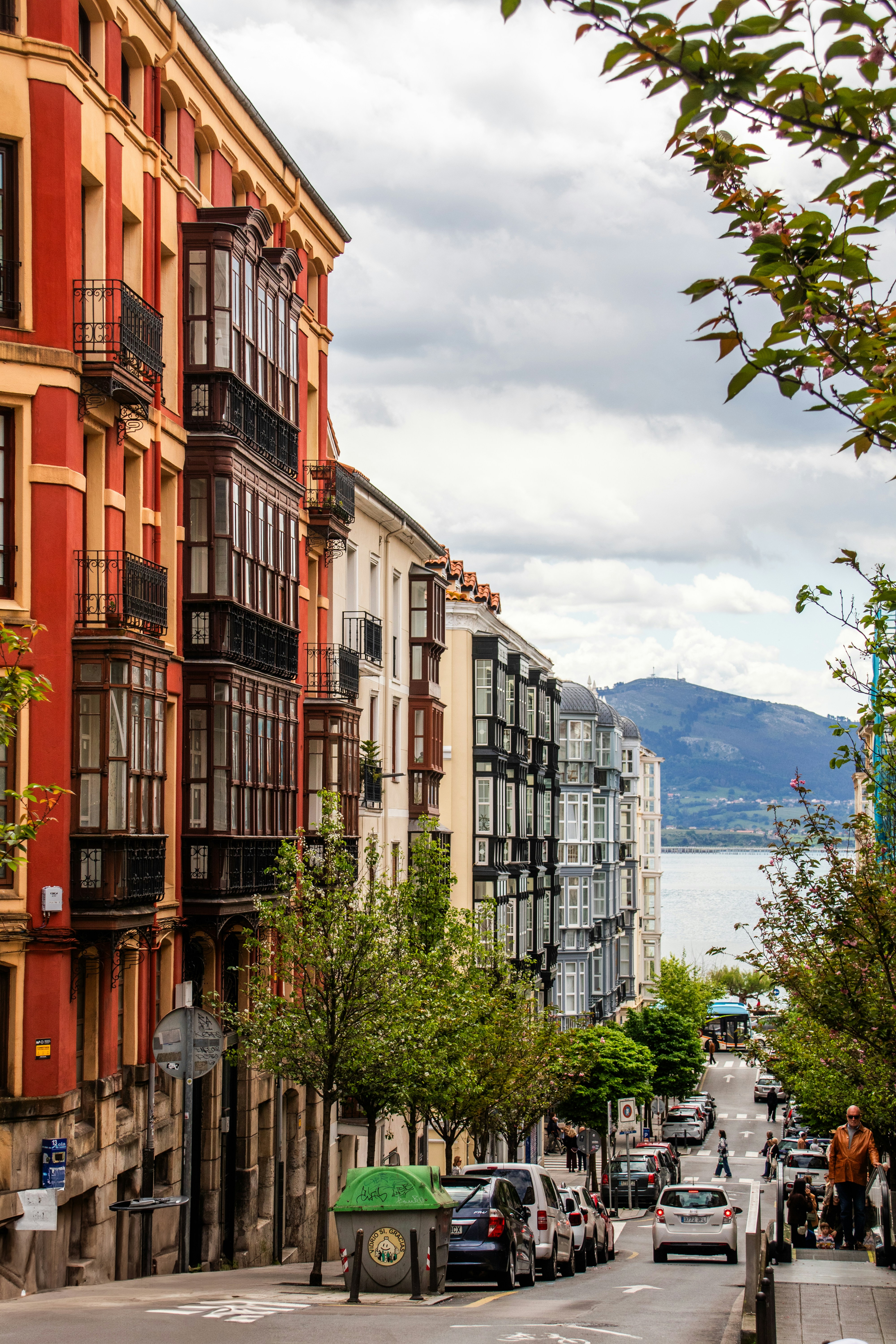 Charming street lined with colorful buildings leading to a serene waterfront view, dotted with trees and vehicles. The scene captures a blend of urban life and nature.