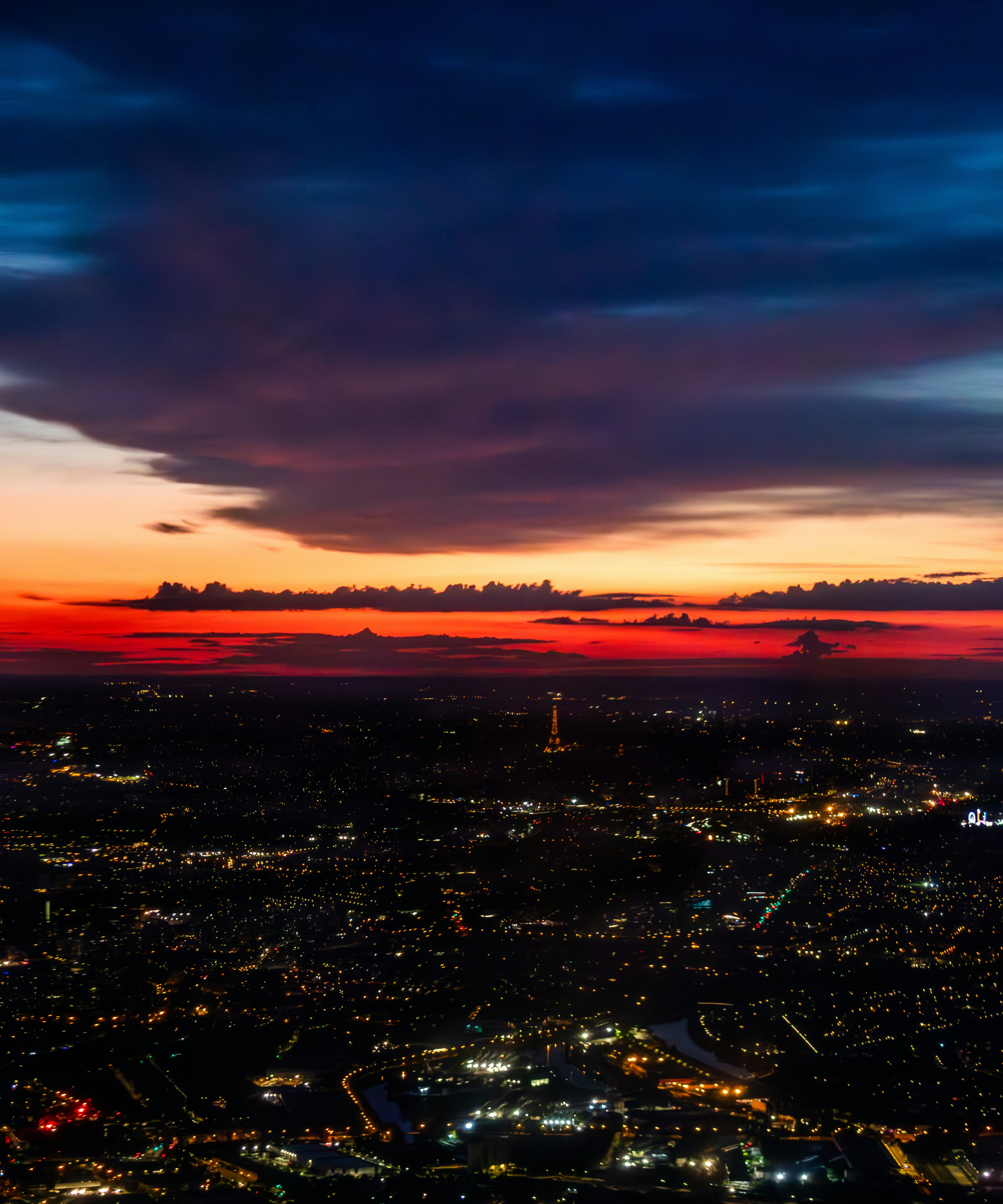Vibrant sunset casting hues over a sprawling cityscape, with distant lights twinkling below. The sky transitions from deep blue to warm orange and red.