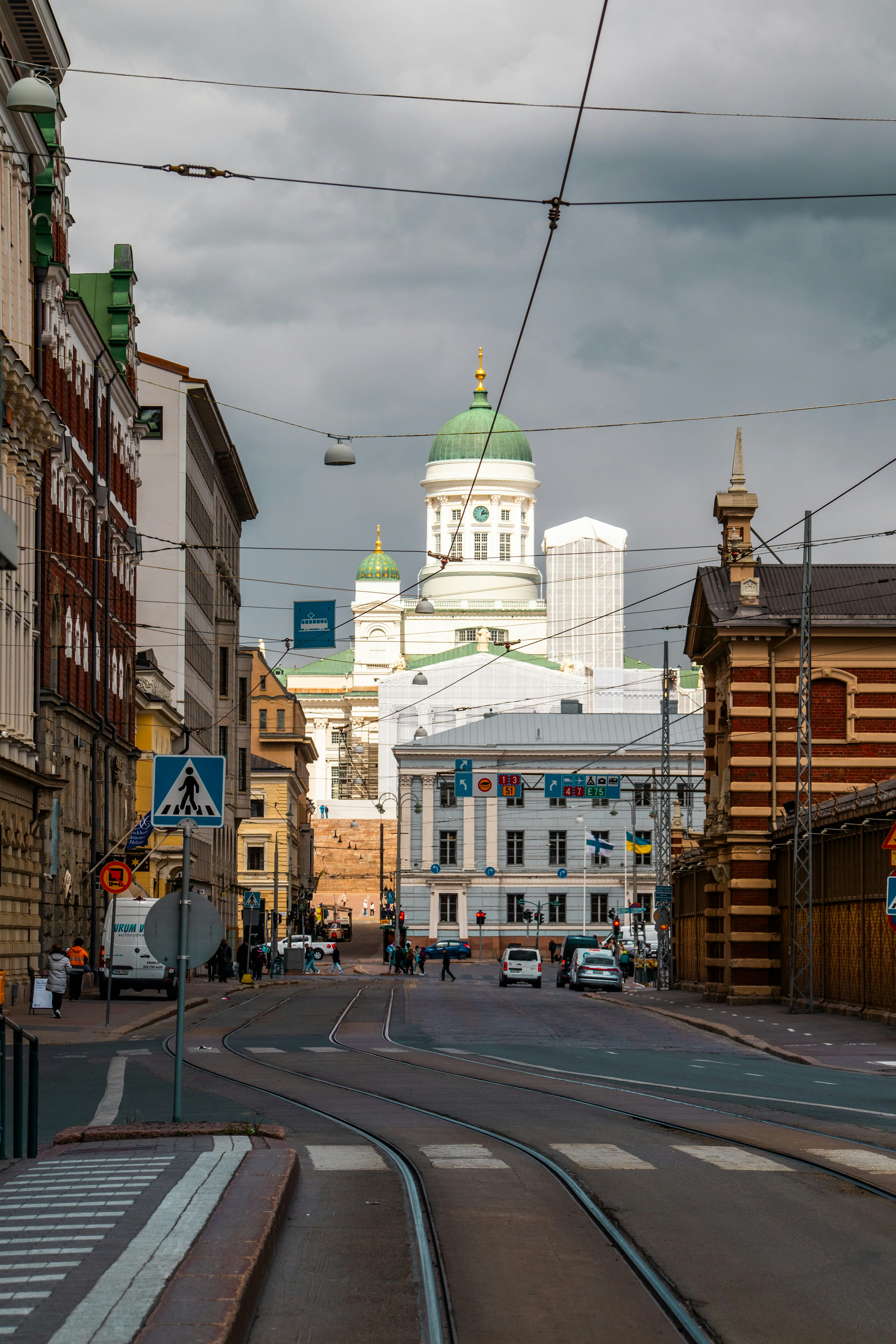 Historic architecture lines a bustling street in Helsinki, with the iconic green dome of a cathedral peeking through the buildings. Streetcars and pedestrians animate the urban landscape.