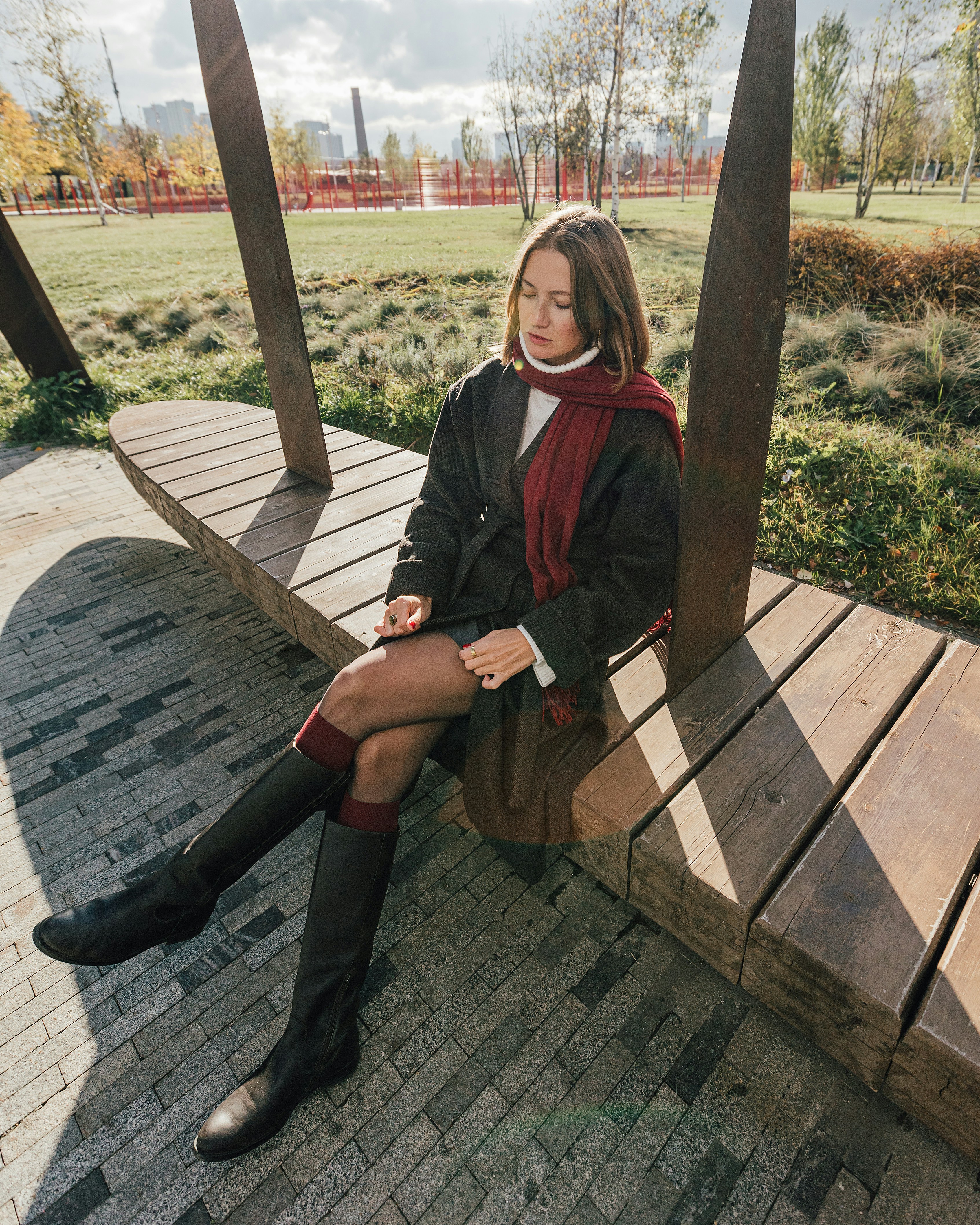 A woman seated on a wooden bench, wrapped in a cozy scarf, enjoying the autumn sun in a park setting.