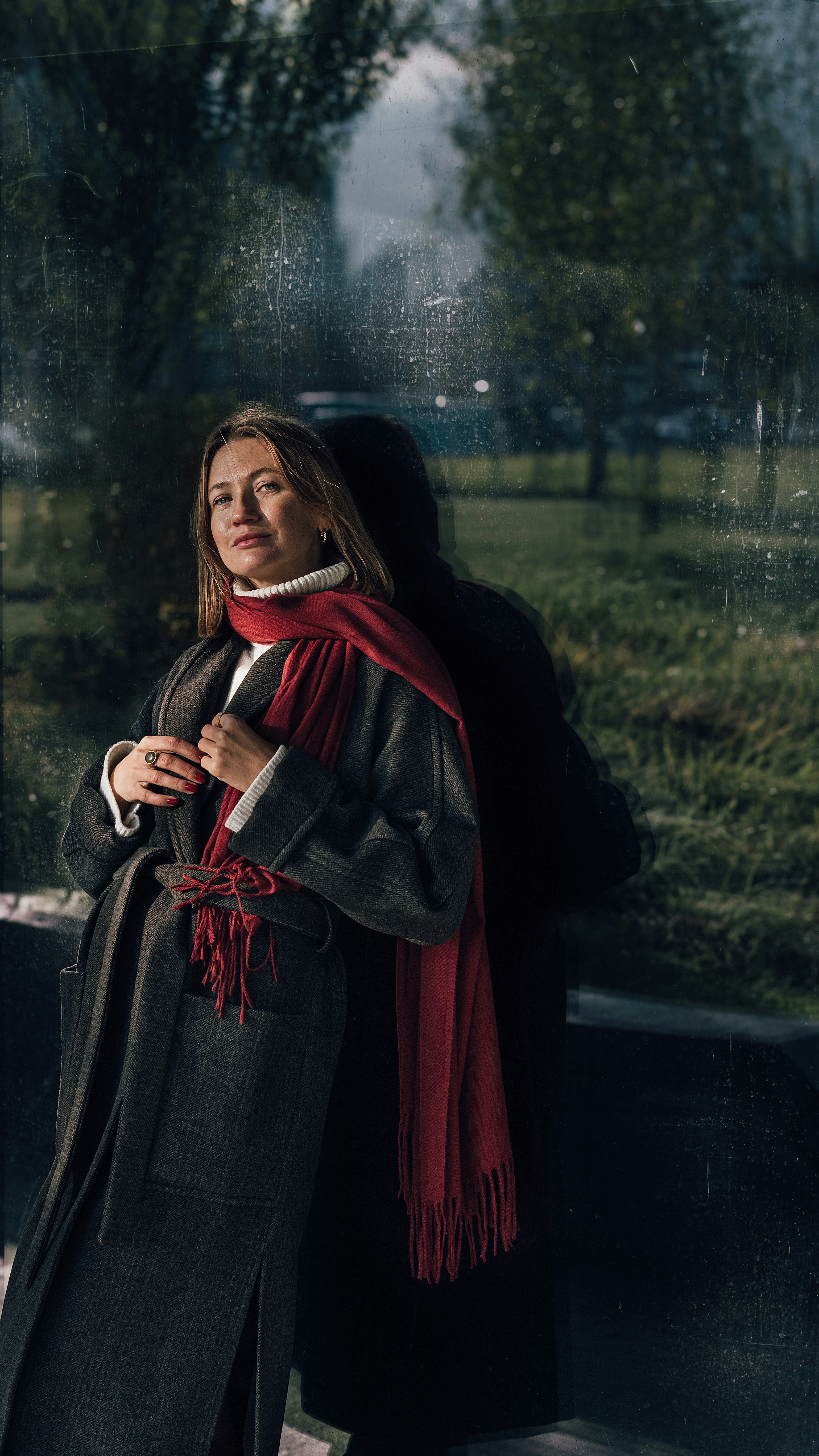A woman in a stylish coat and red scarf stands against a backdrop of greenery, casting a soft shadow. The interplay of light and texture enhances the scene's depth.