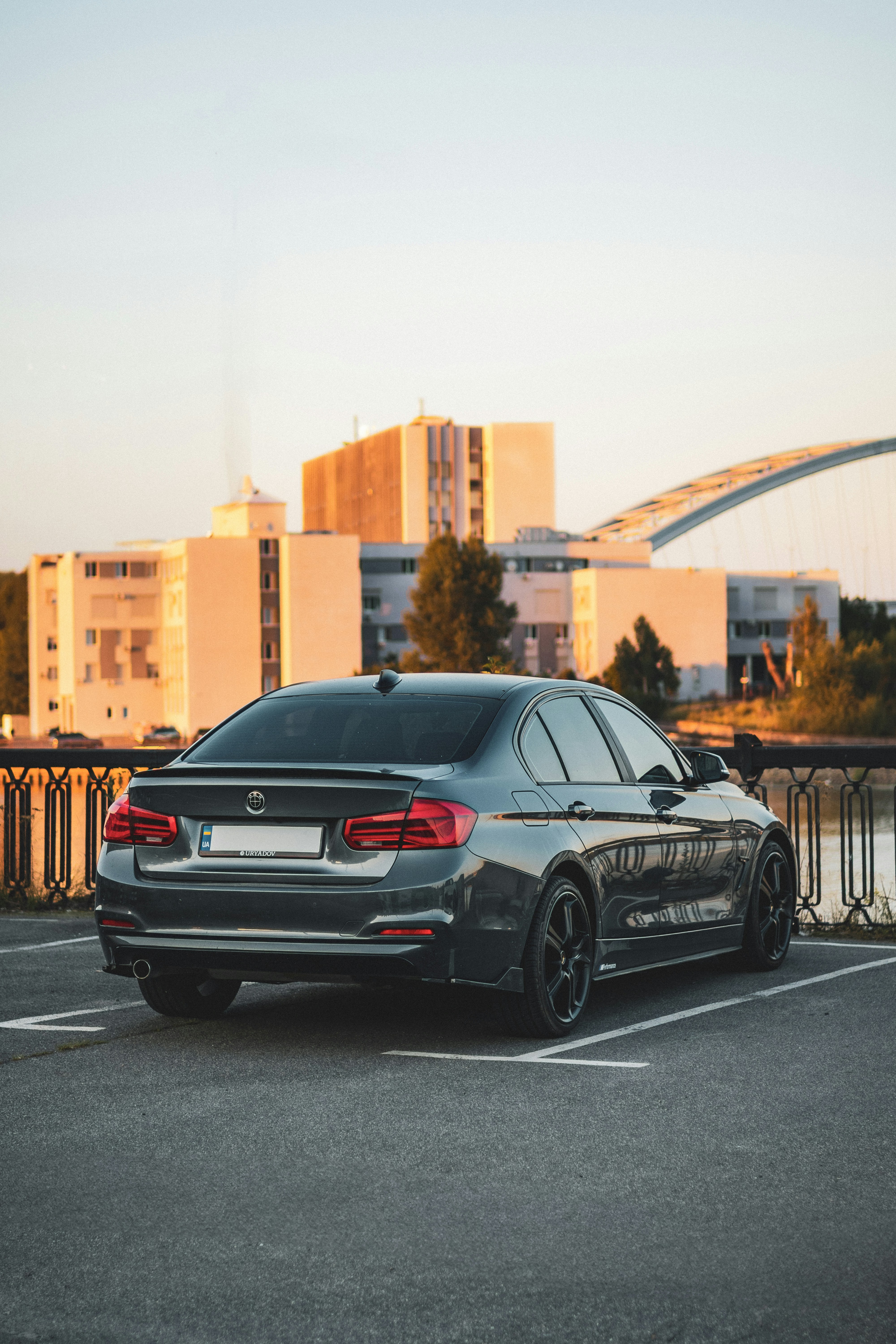A sleek gray sedan parked by the riverside at sunset, framed by modern architecture and a bridge in the background.