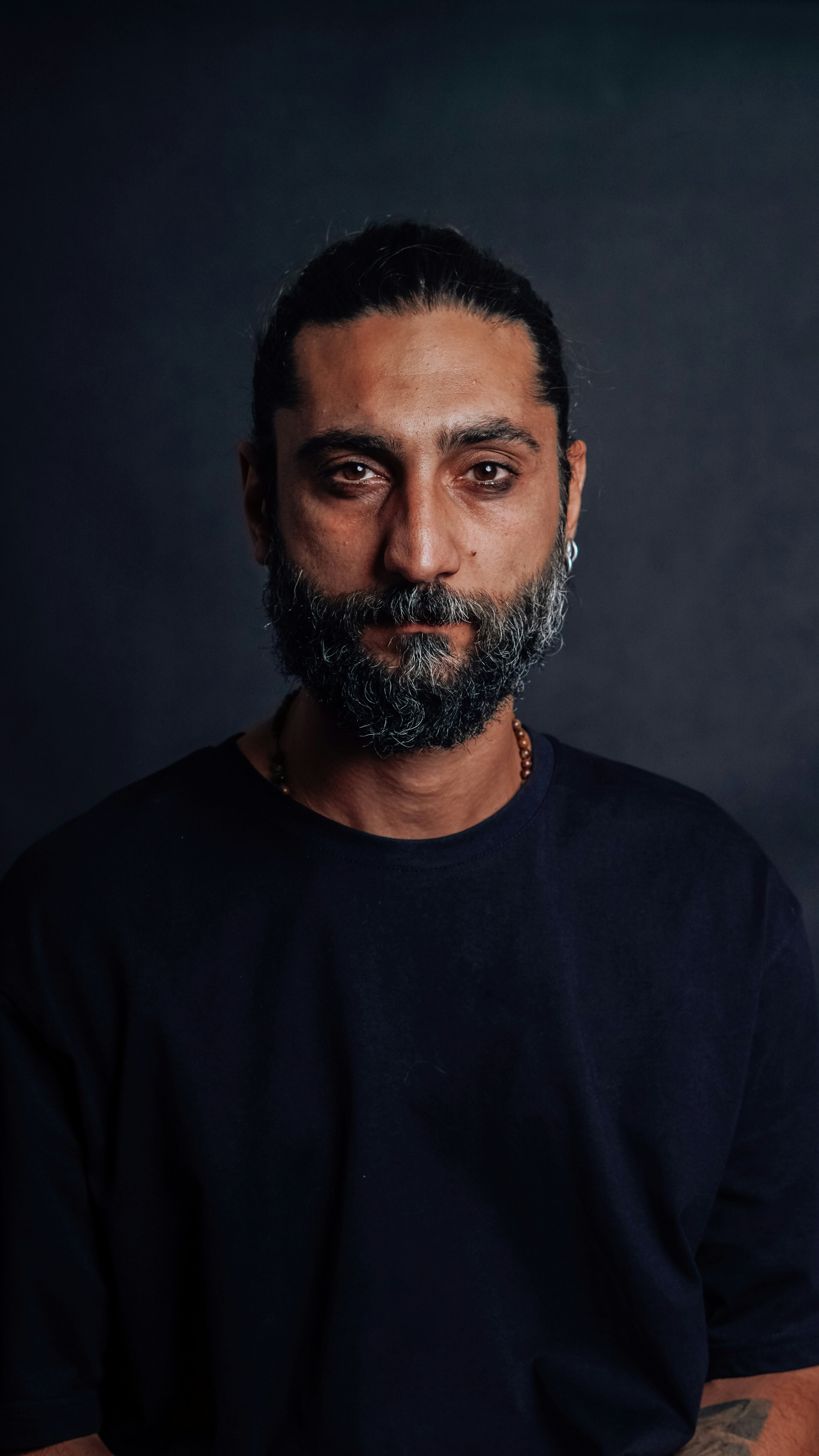 A high-resolution portrait of a bearded man wearing a casual t-shirt and a single earring, set against a dark background. Subtle, moody lighting emphasizes his facial hair, accessories, and modern masculine style — ideal for editorial, lifestyle, and fashion projects. | A man with a beard poses for a portrait.