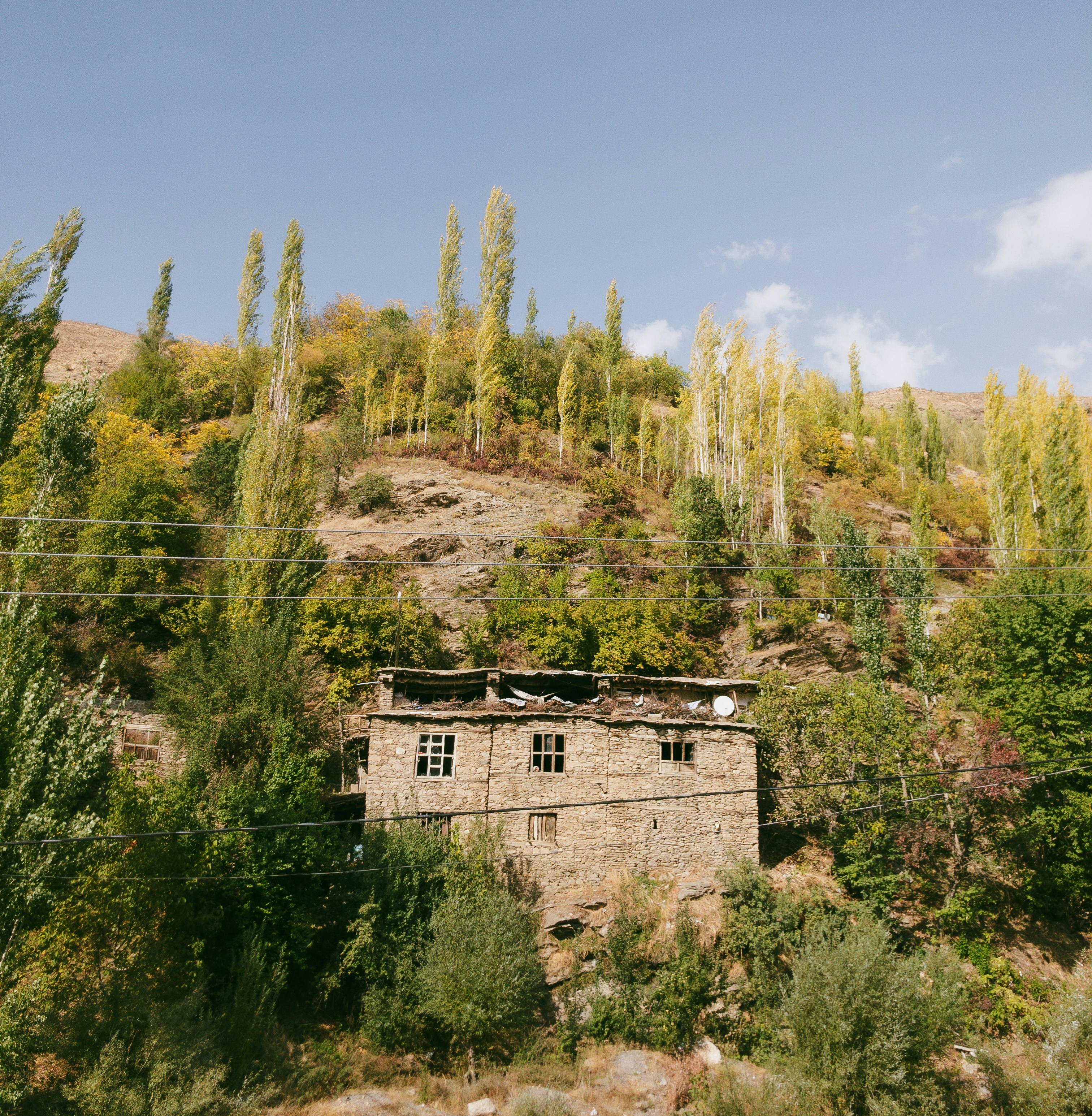 Weathered stone house nestled among vibrant autumn foliage on a hillside, with power lines tracing the landscape.