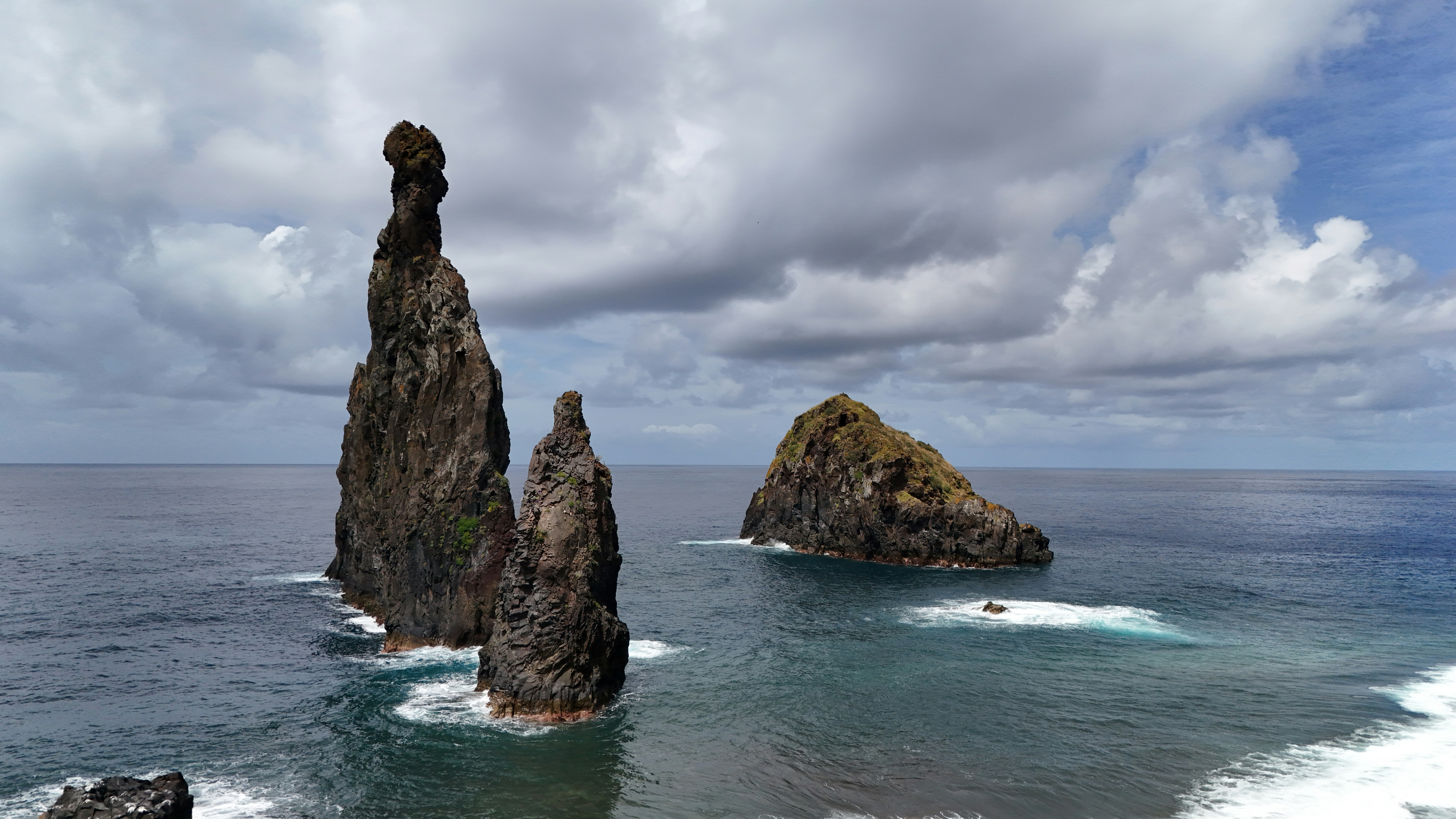 Three towering rock formations rise from the ocean, surrounded by gentle waves and a dramatic sky. The scene captures the raw beauty of coastal geology.