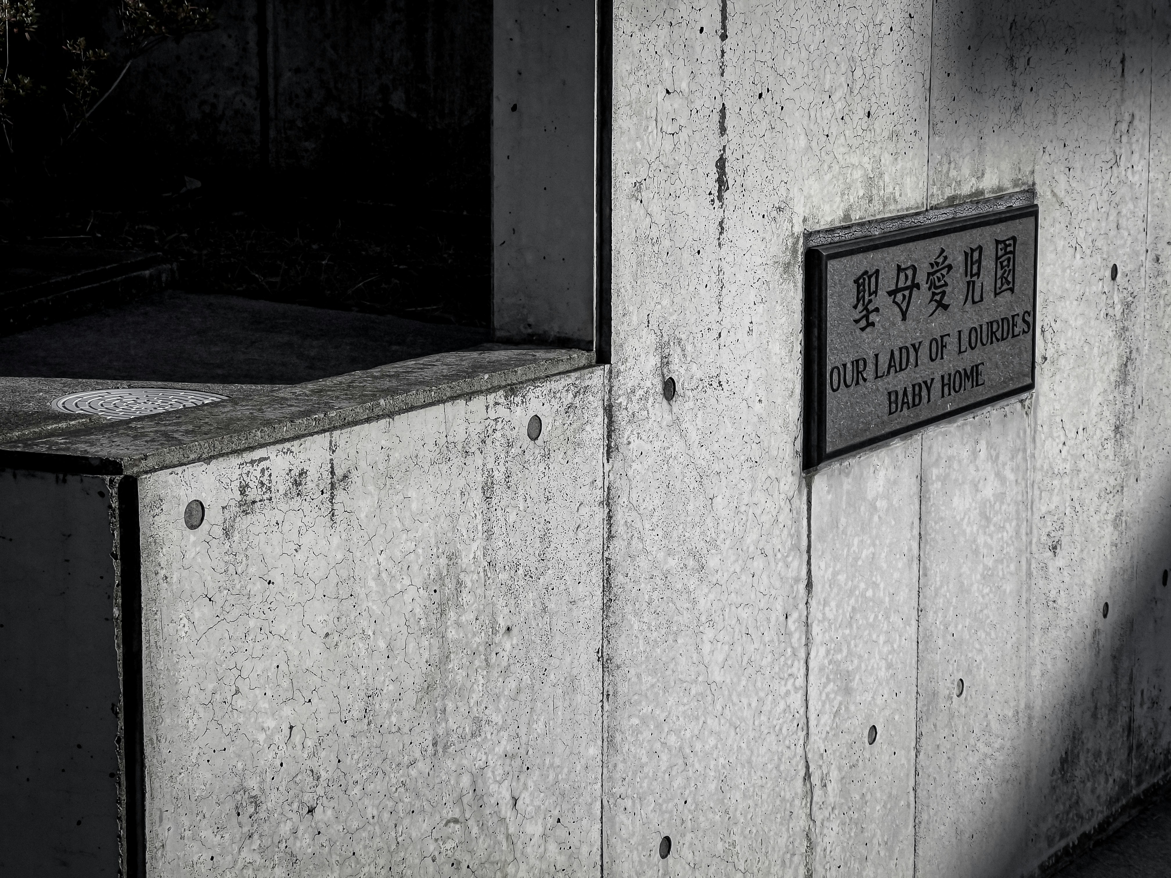Concrete wall with a plaque reading 'Our Lady of Lourdes Baby Home' alongside a shadowed alcove.