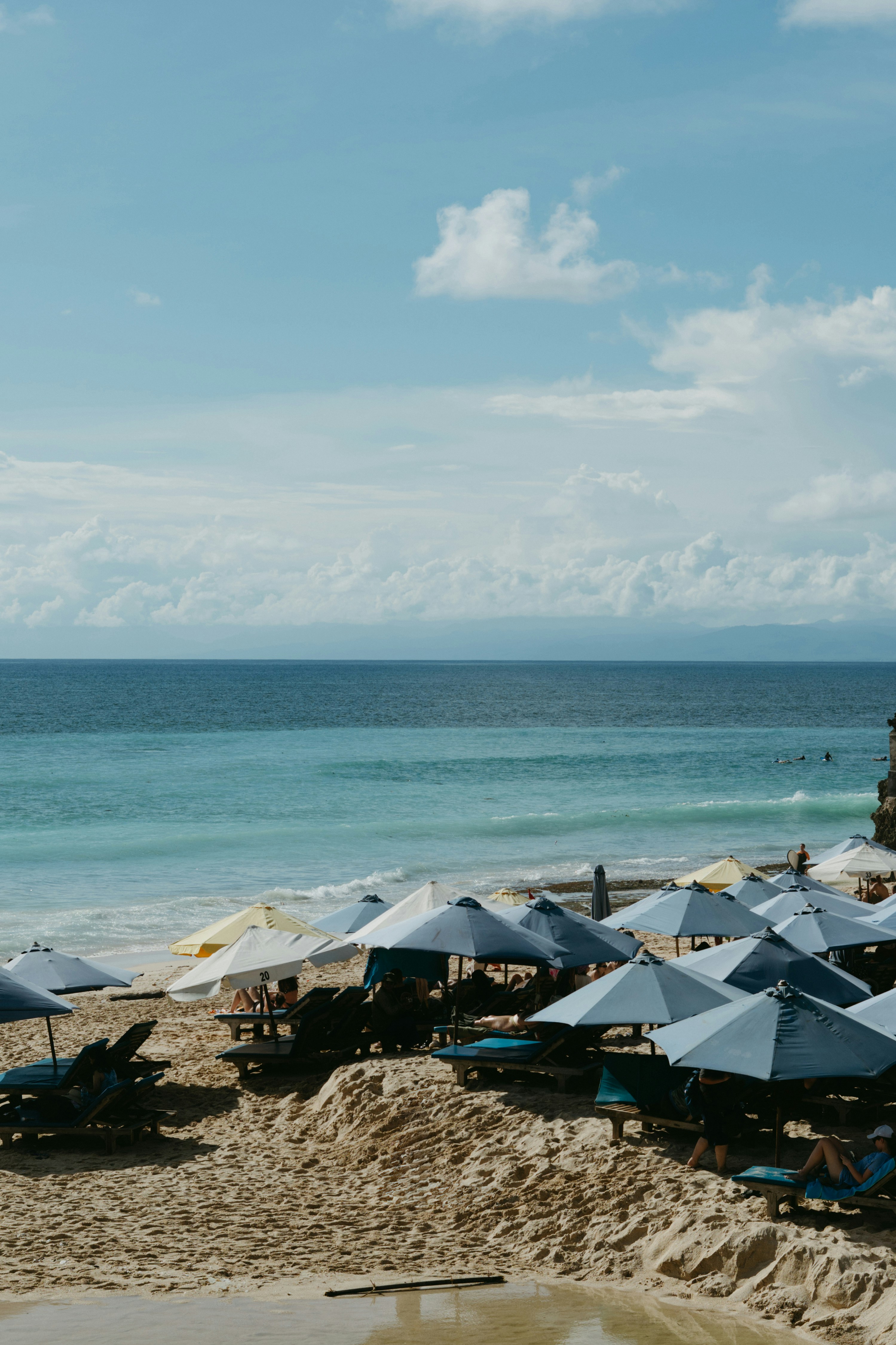 Beach umbrellas providing shade to sunbathers on a sandy shore, with gentle waves lapping at the coastline under a clear blue sky.