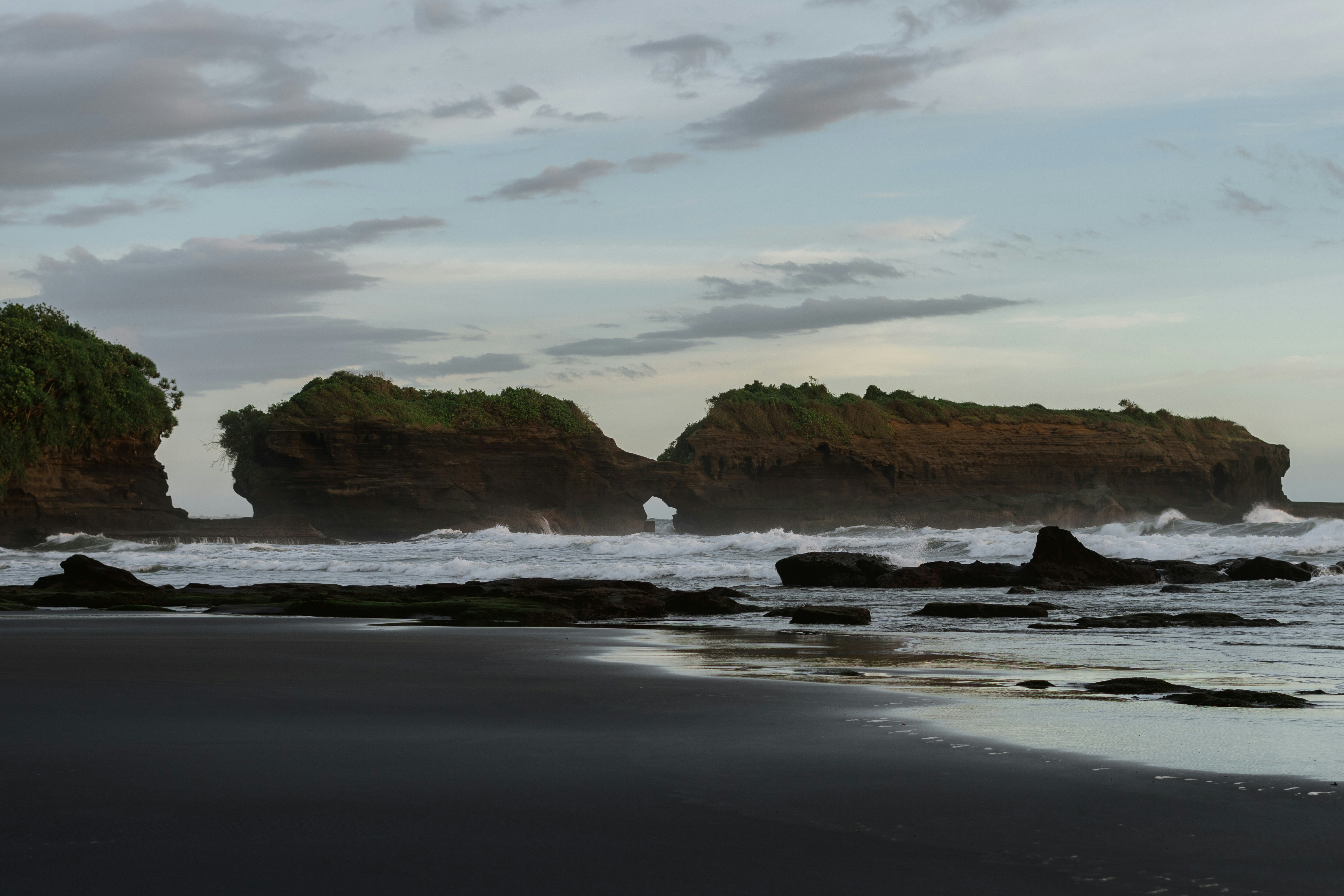 Twin rock formations adorned with lush greenery emerge from the waves, creating a natural archway against the horizon. The tranquil beach reflects the soft hues of the sky.