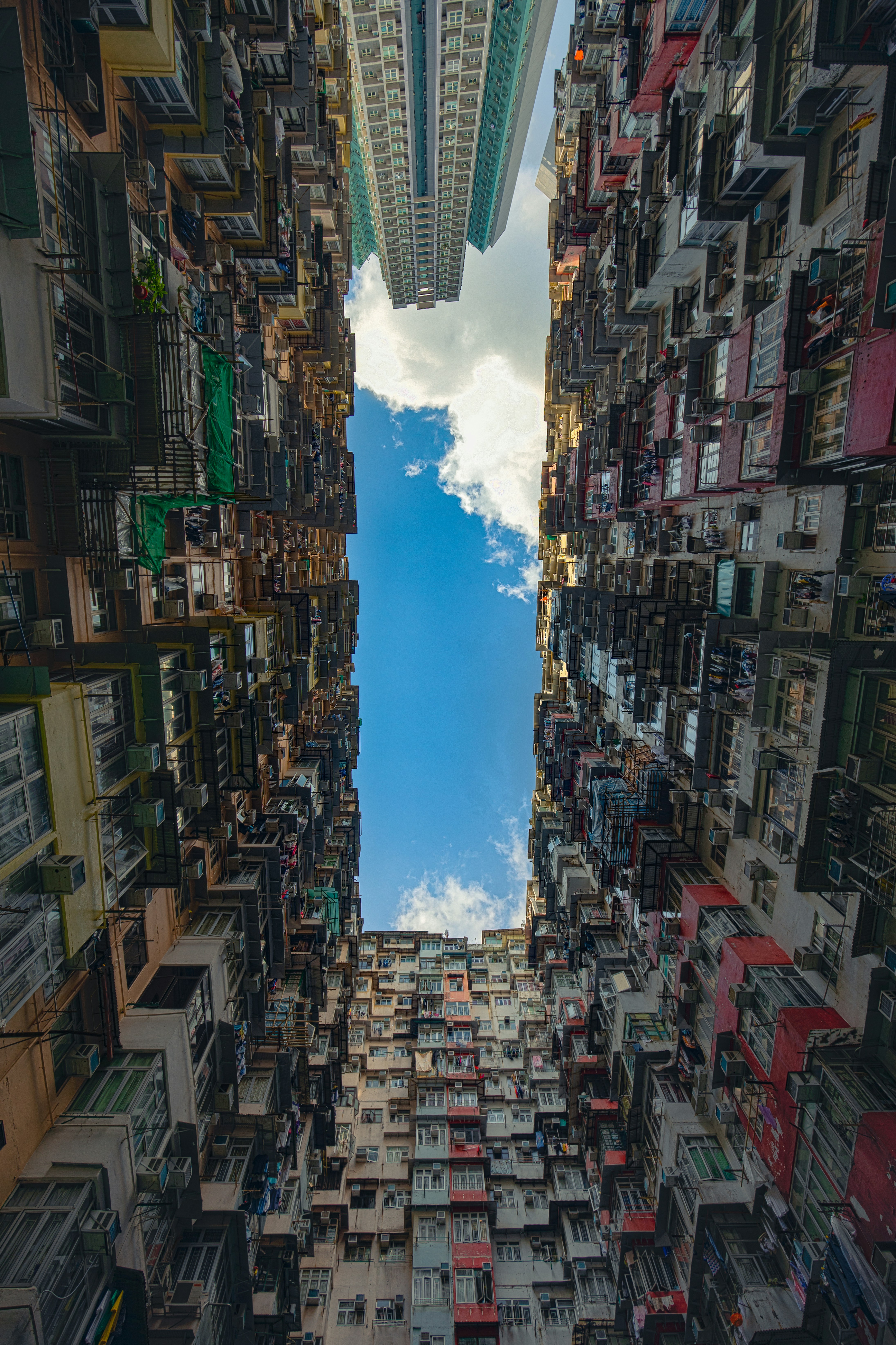 Looking up through towering apartment buildings in Hong Kong, revealing a patch of blue sky framed by dense urban architecture. | Looking up at packed hong kong buildings.