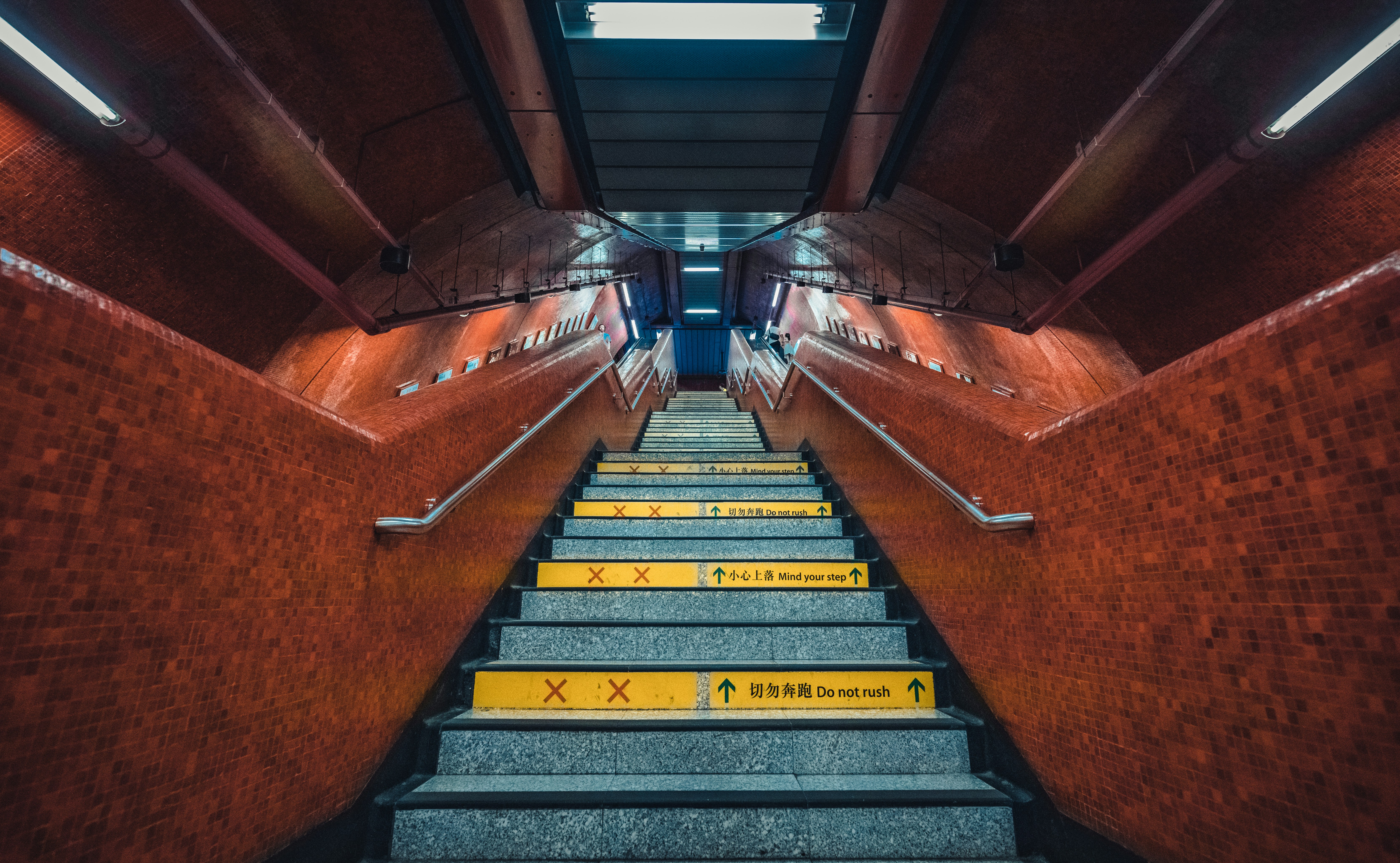 A symmetrical view of a subway stairway in Hong Kong, framed by red-tiled walls and fluorescent lights, leading upward toward the city above. | Staircase leads upwards through a red tunnel.