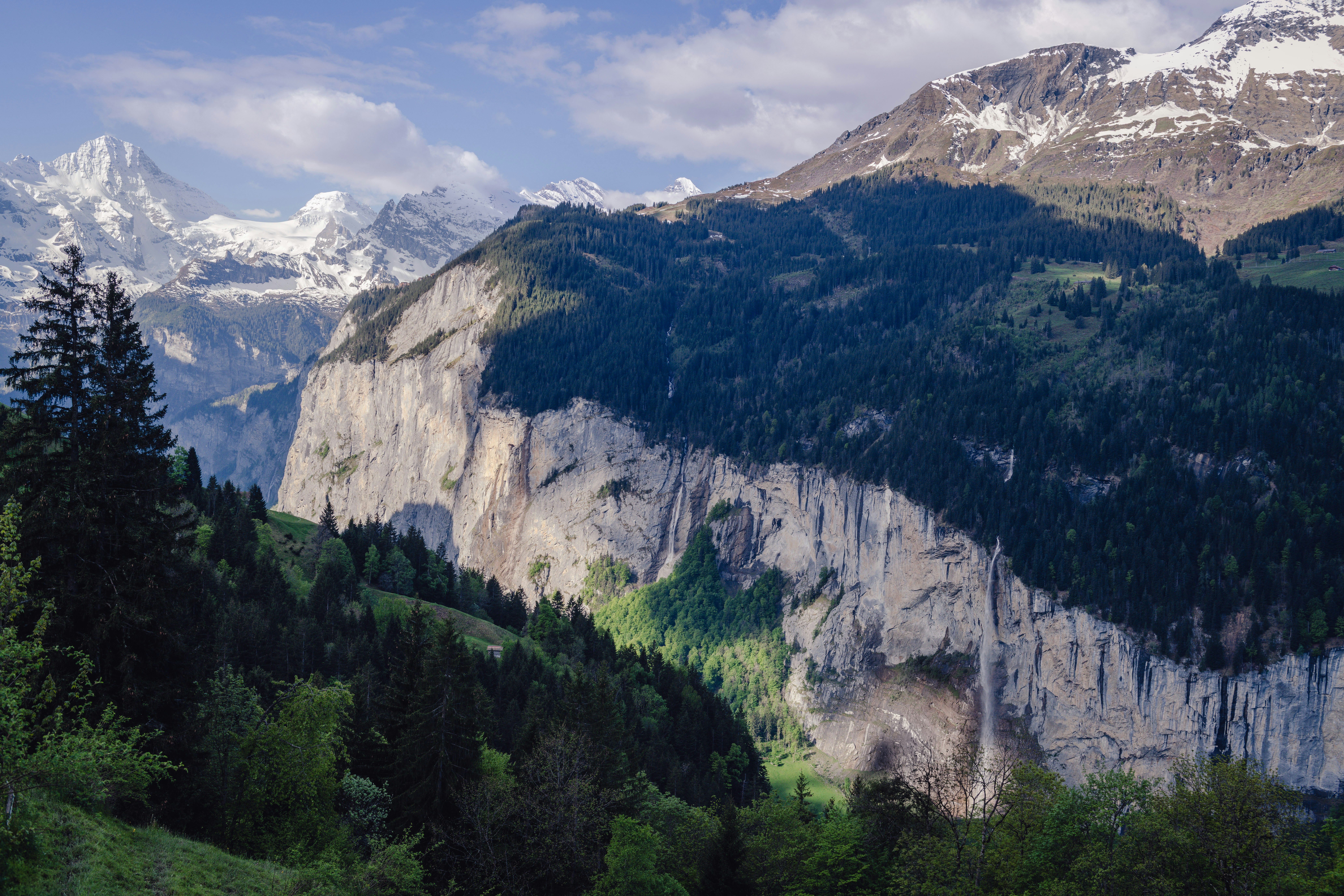 Towering cliffs and lush greenery juxtapose against snow-capped peaks under a dynamic sky.