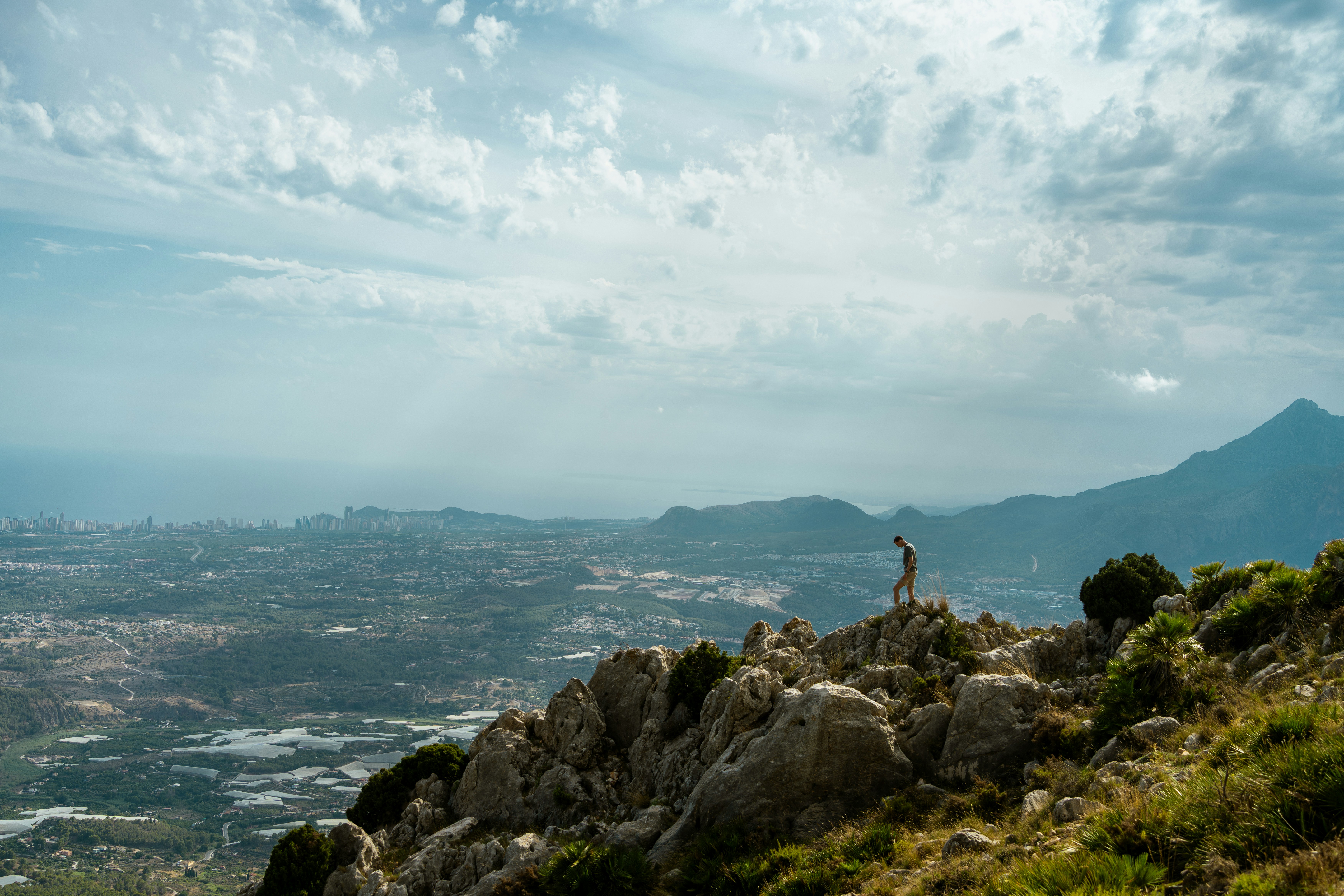 A lone figure stands atop a rocky summit, gazing over a sprawling landscape that meets the sea. The interplay of light and shadow creates a dramatic backdrop.