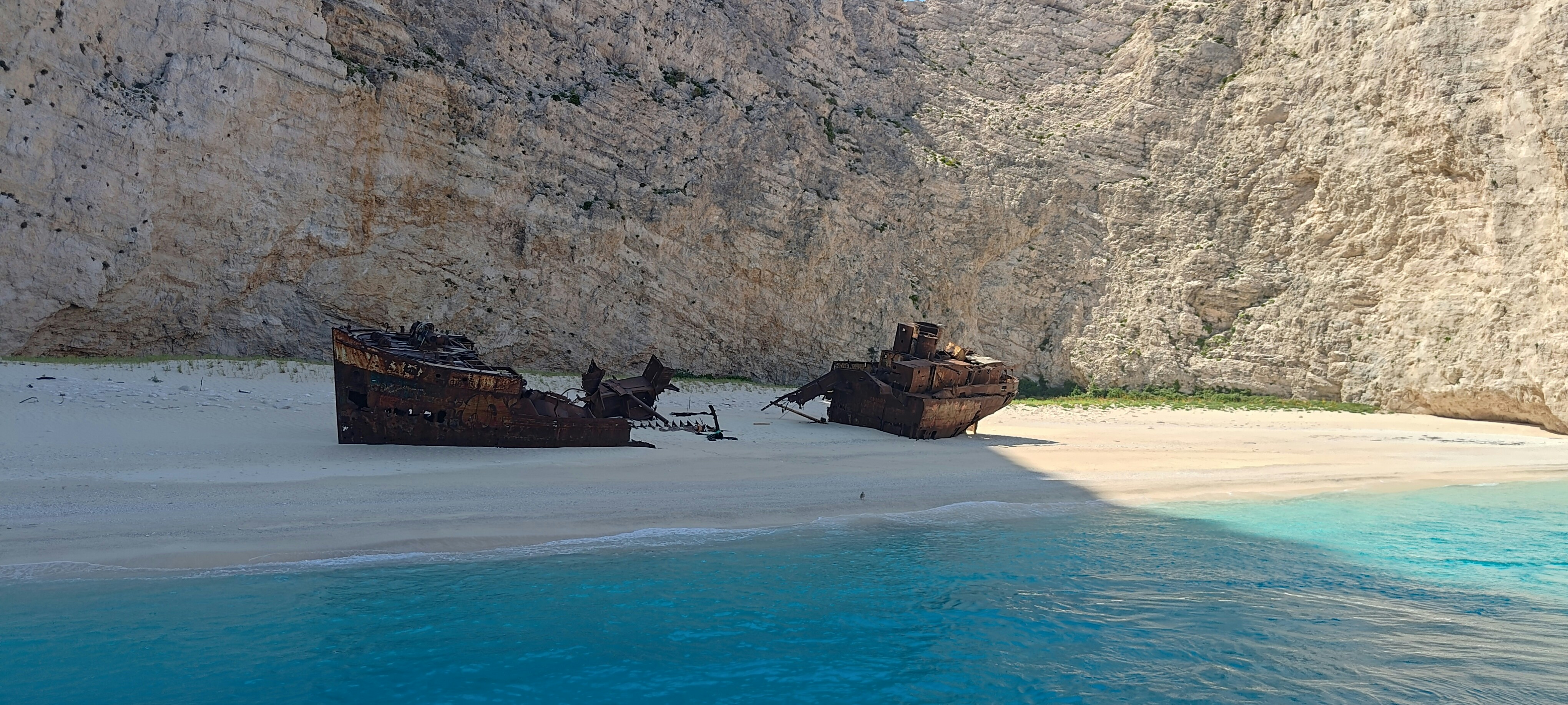 The famous shipwreck in Navaggio harbour, Zakynthos, Greece. | Shipwrecks sit on a sandy beach, next to the ocean.