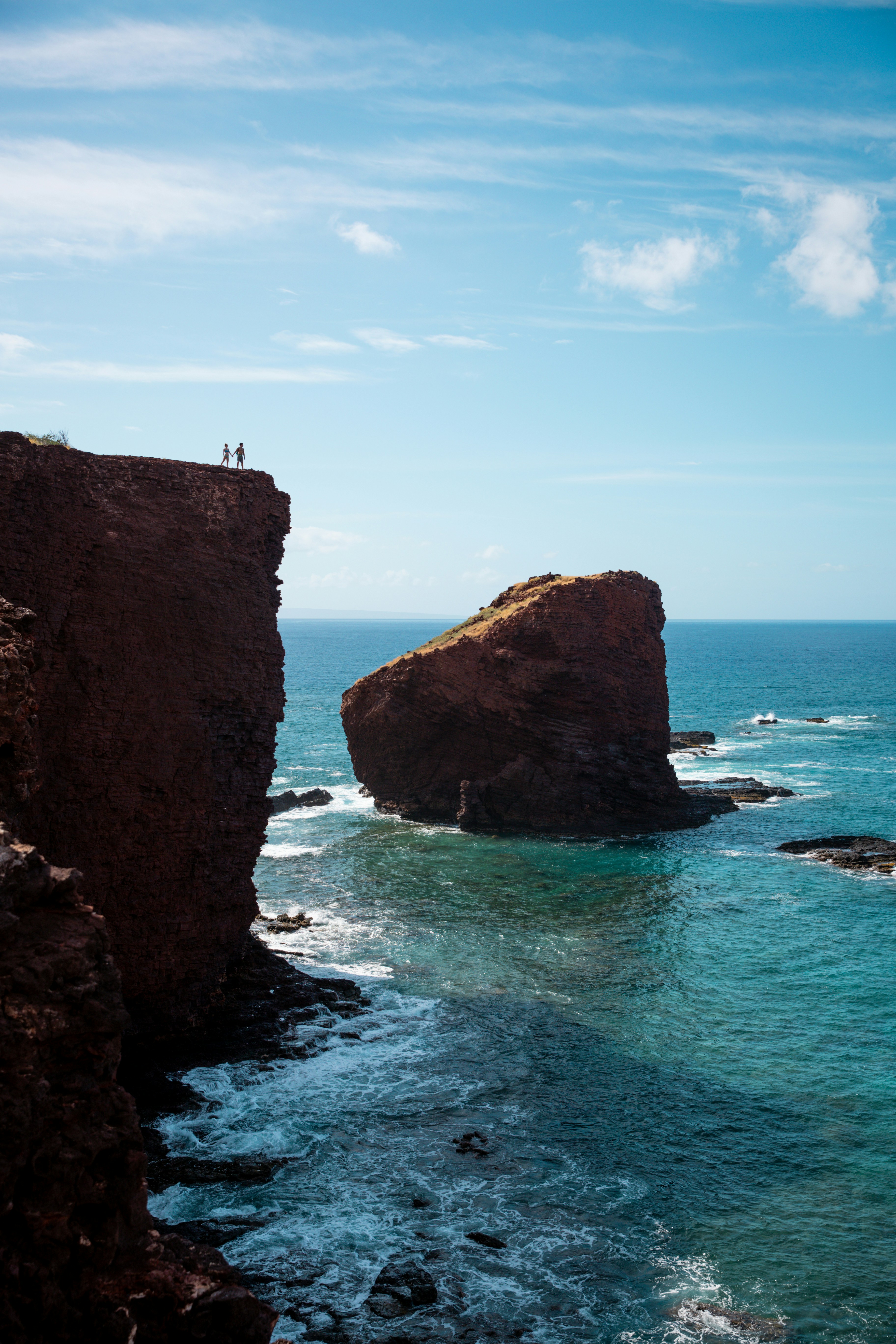 Two towering rock formations rise from the turquoise waters, with a pair of figures silhouetted atop the cliff, capturing the essence of adventure and exploration.