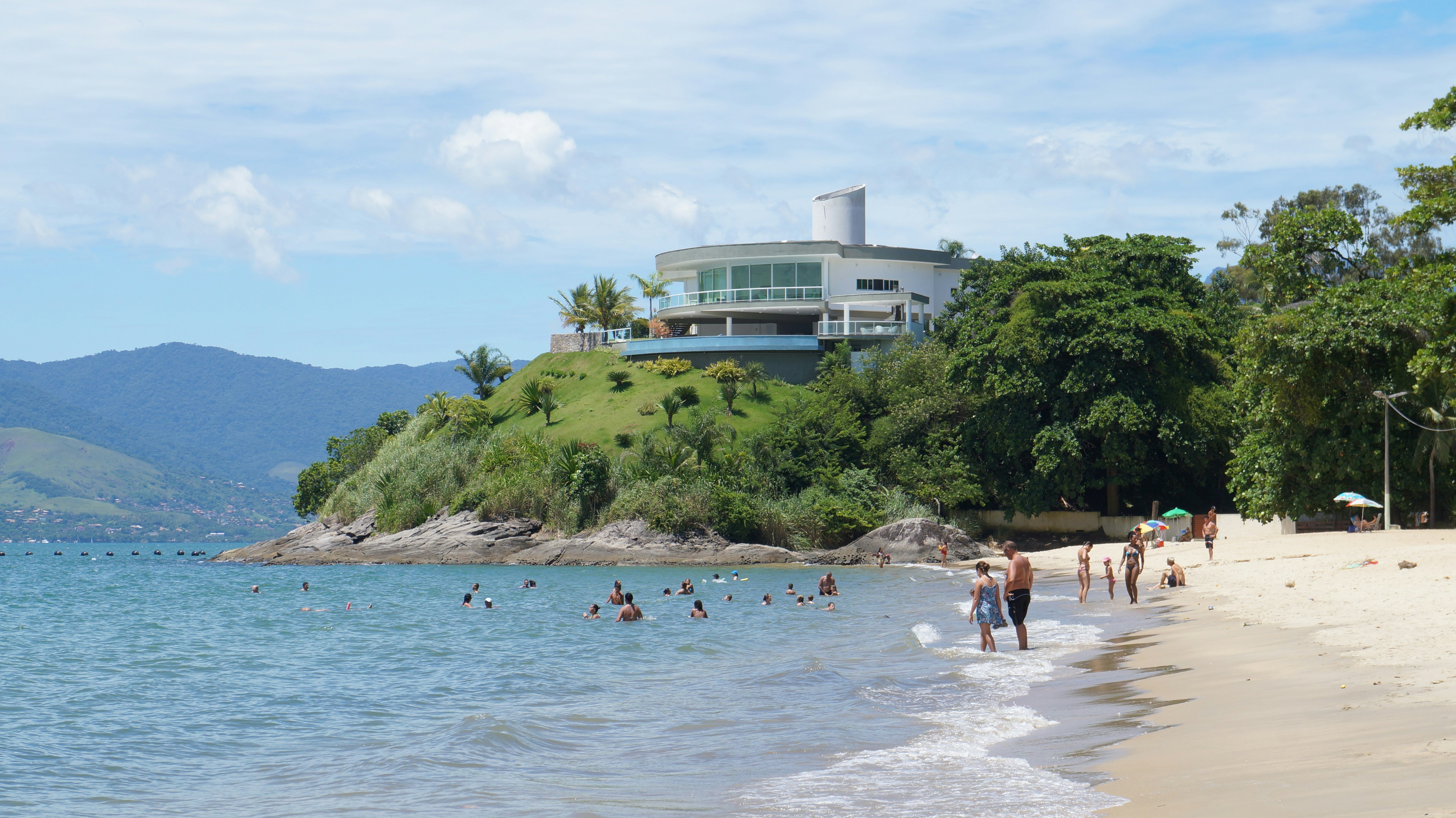 A contemporary house perched on a lush hill overlooking a sandy beach, with people enjoying the sun and water. The scene captures a tranquil coastal atmosphere.