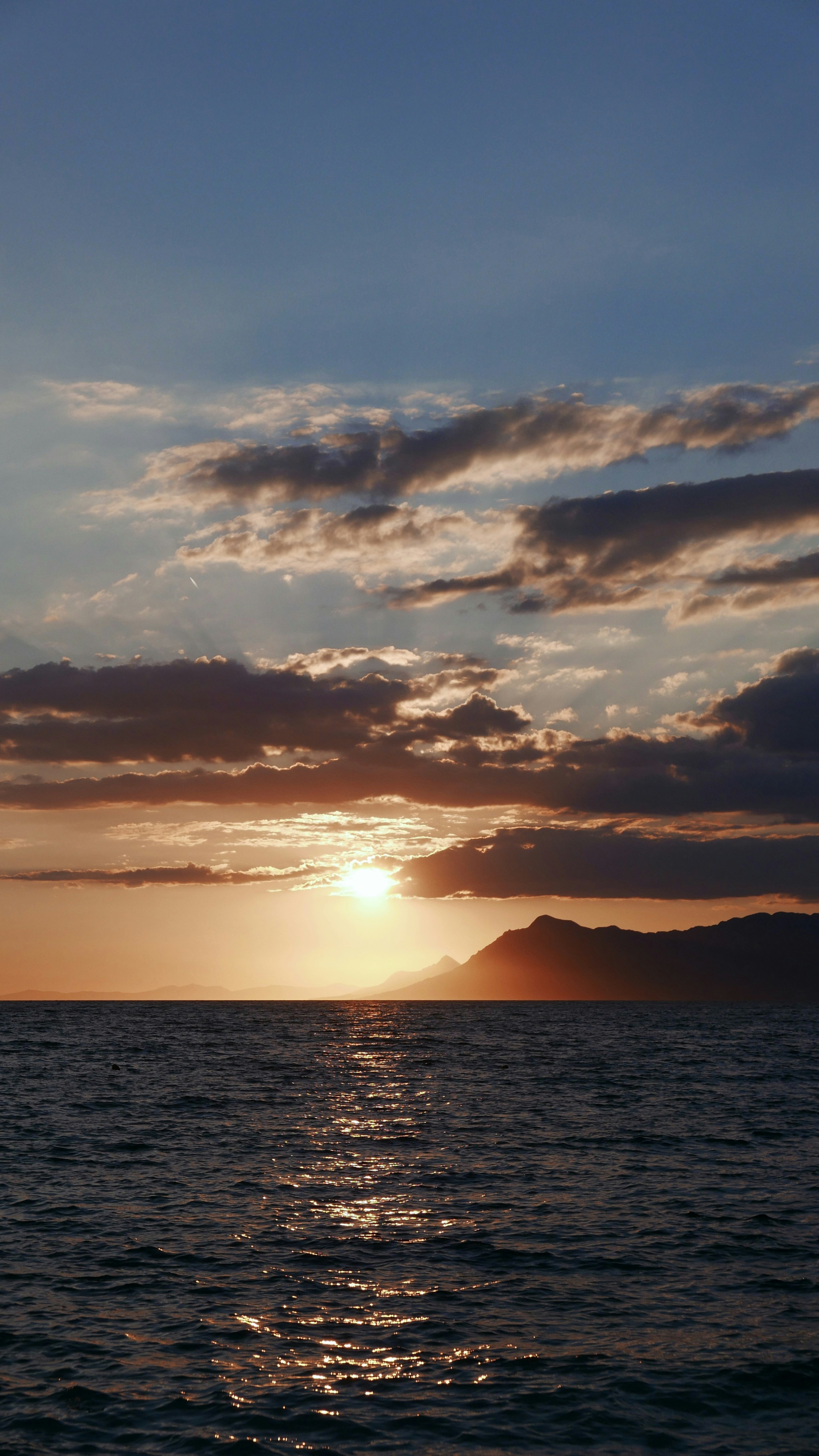 Golden sun setting behind distant mountains, casting shimmering reflections on the ocean surface. Soft clouds drift across the sky, enhancing the tranquil atmosphere.