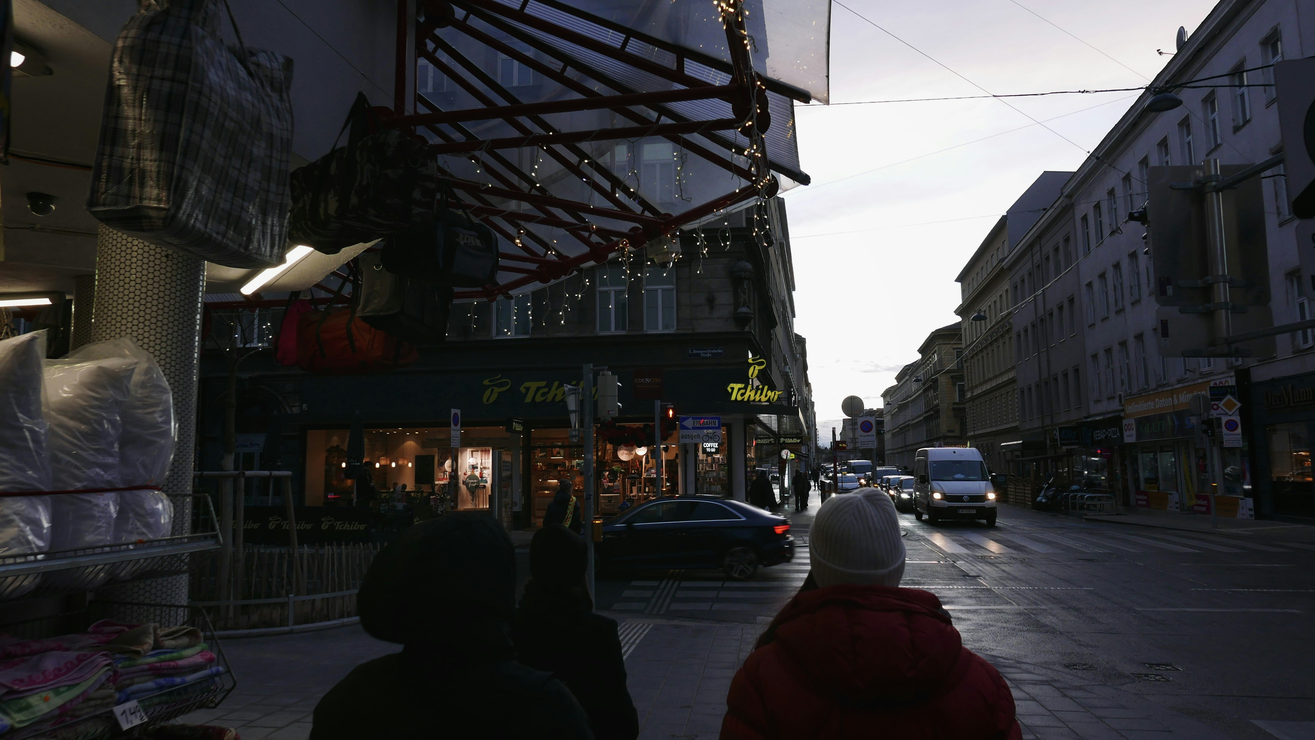 Three figures walk along a city street adorned with festive lights, as evening descends and storefronts glow invitingly.