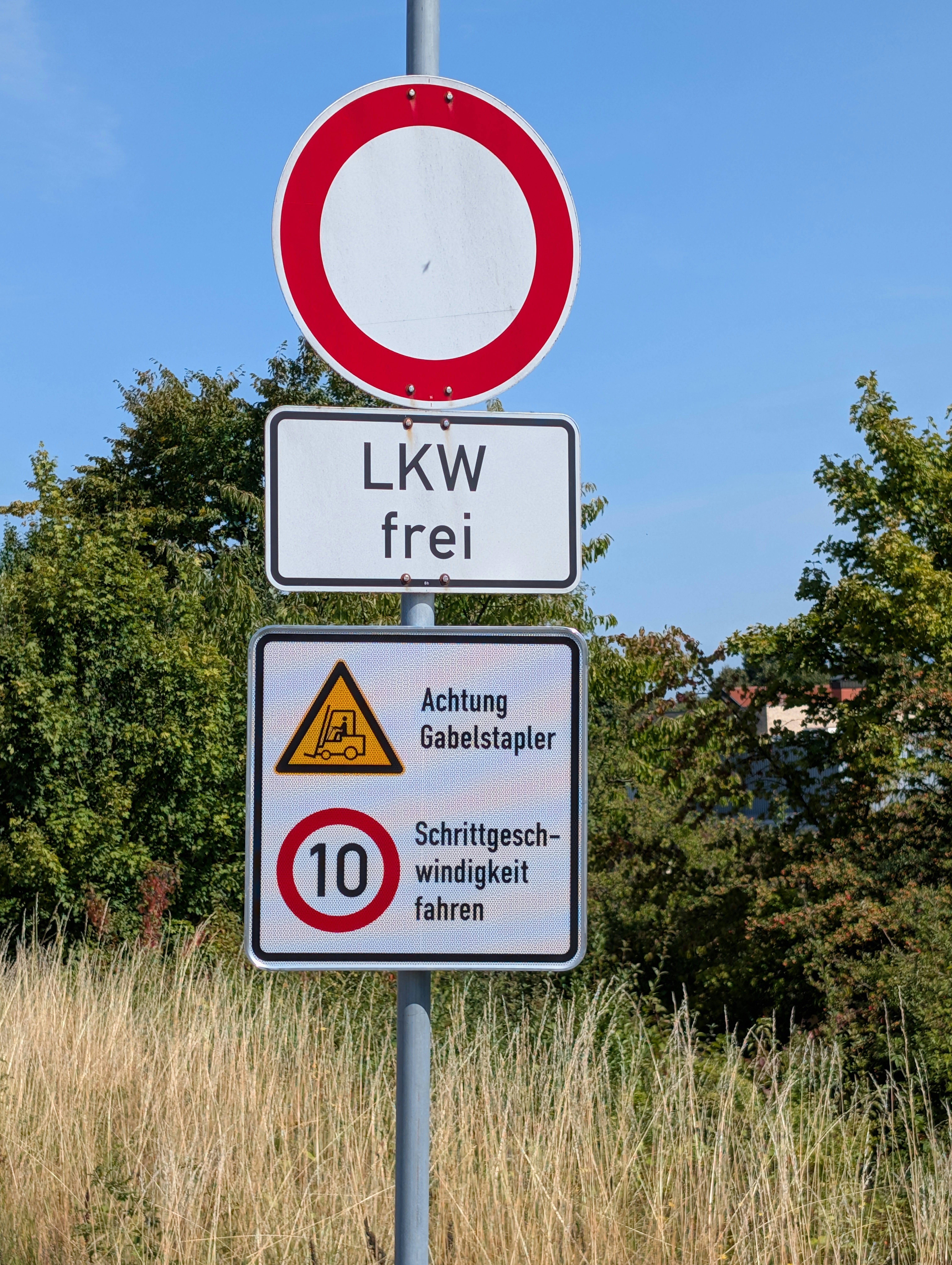 Traffic sign indicating a restricted area for trucks, with additional warnings for forklift operation and speed limits. The scene is set against a backdrop of greenery.