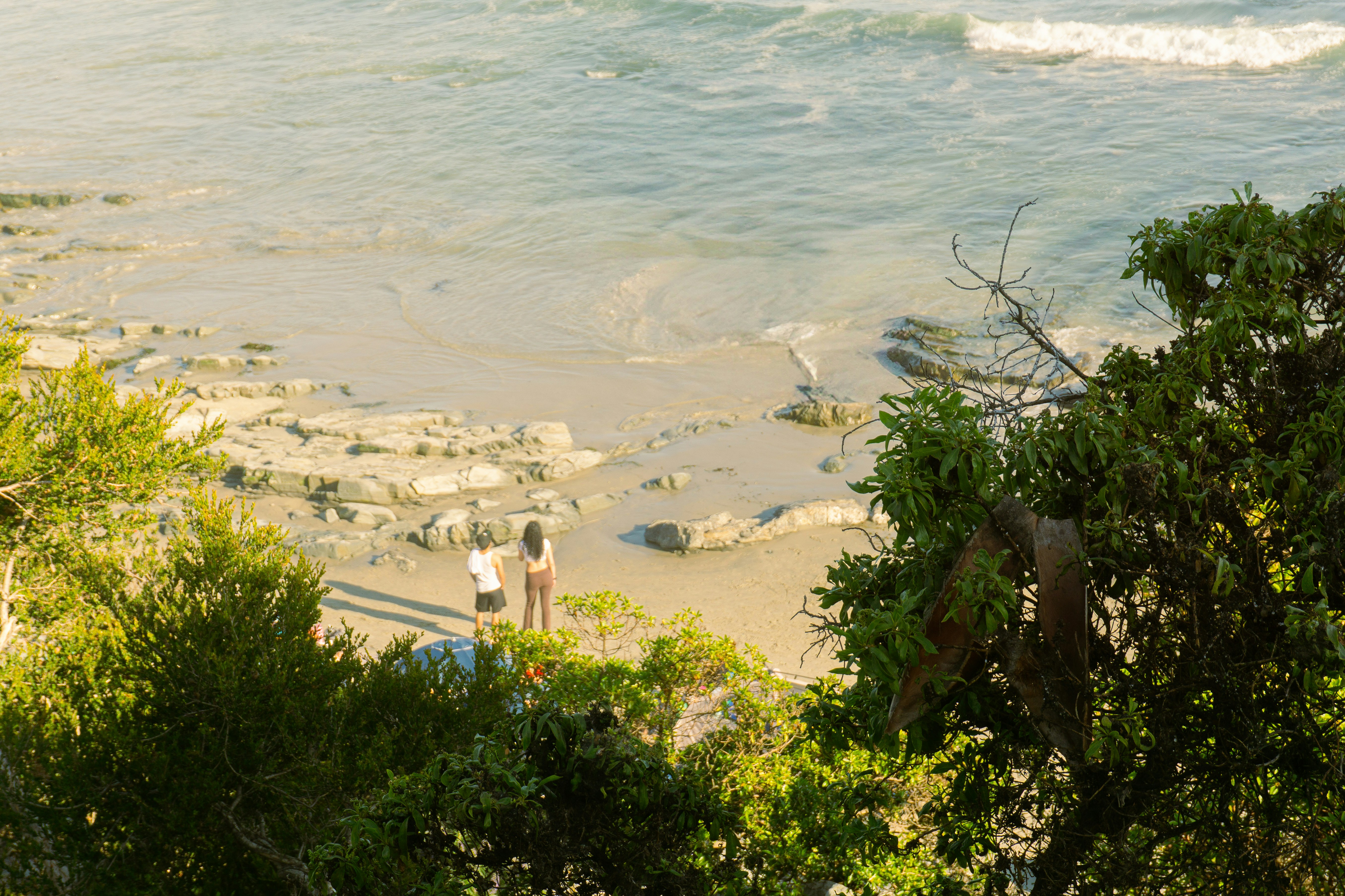 Two figures standing on a sandy beach, framed by lush greenery, with gentle waves lapping at the shore.