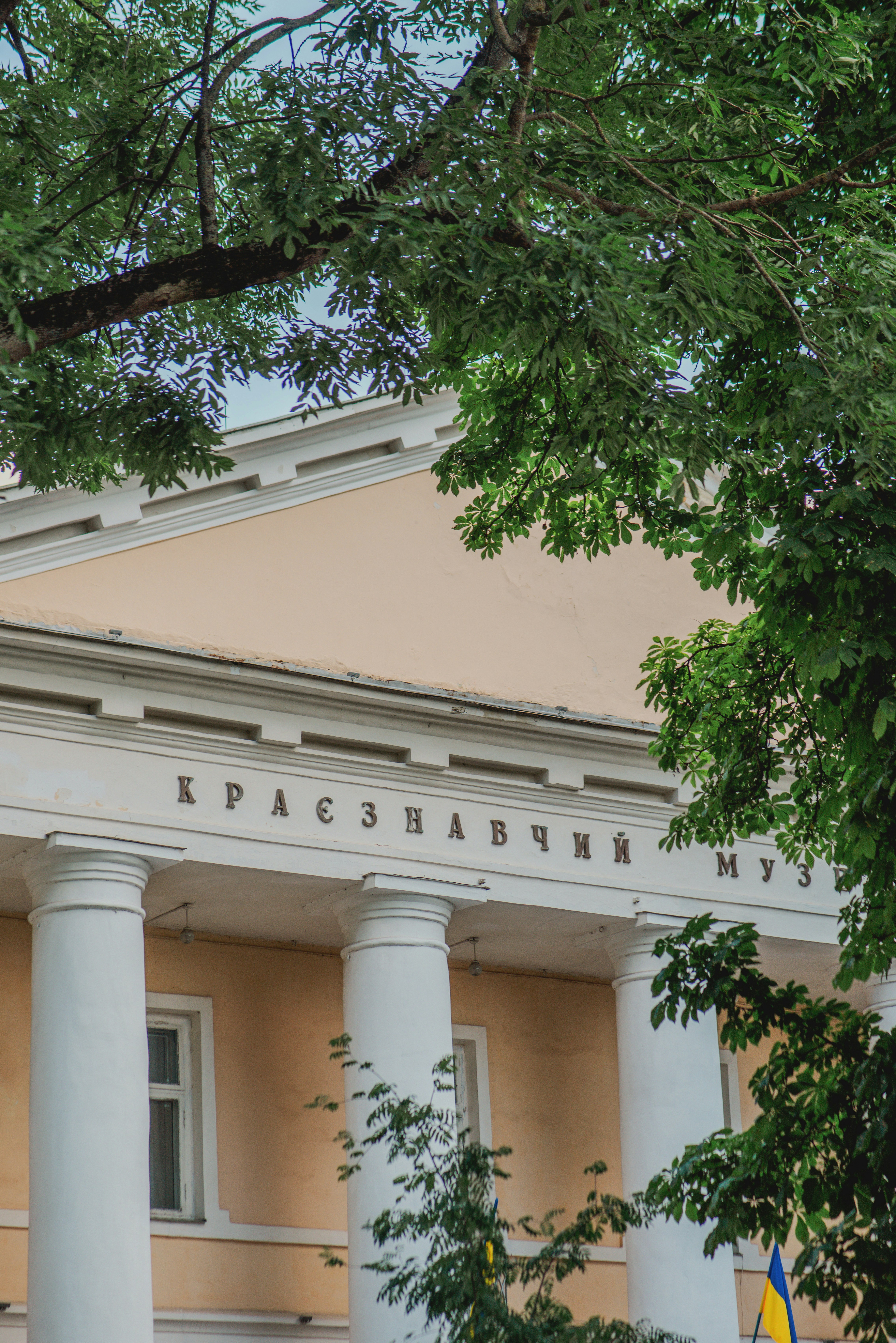 Historic museum facade framed by lush green foliage, showcasing classical architecture and a hint of vibrant color from the flag.