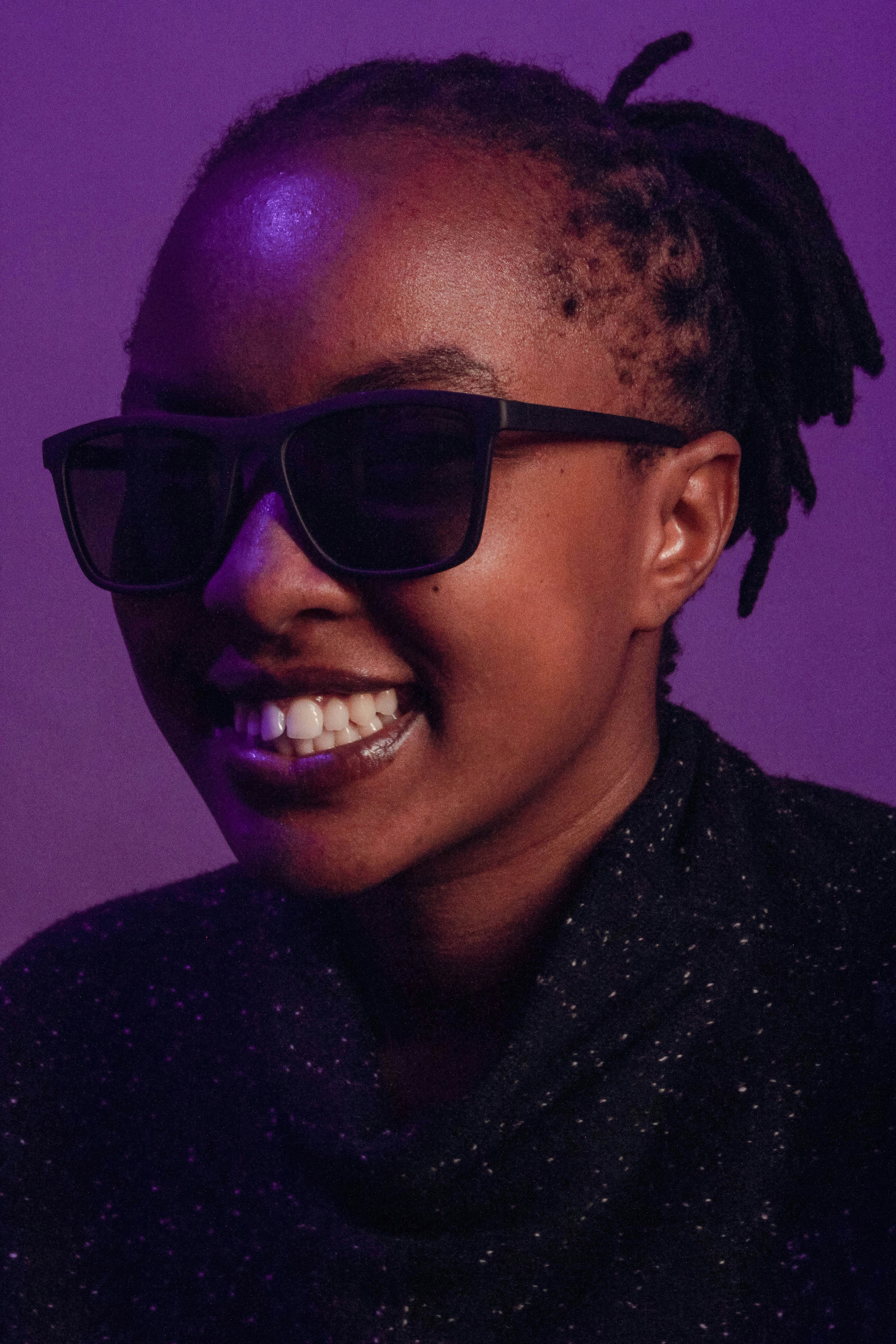 Smiling individual wearing sunglasses, illuminated by soft purple lighting, showcasing a playful and confident demeanor.