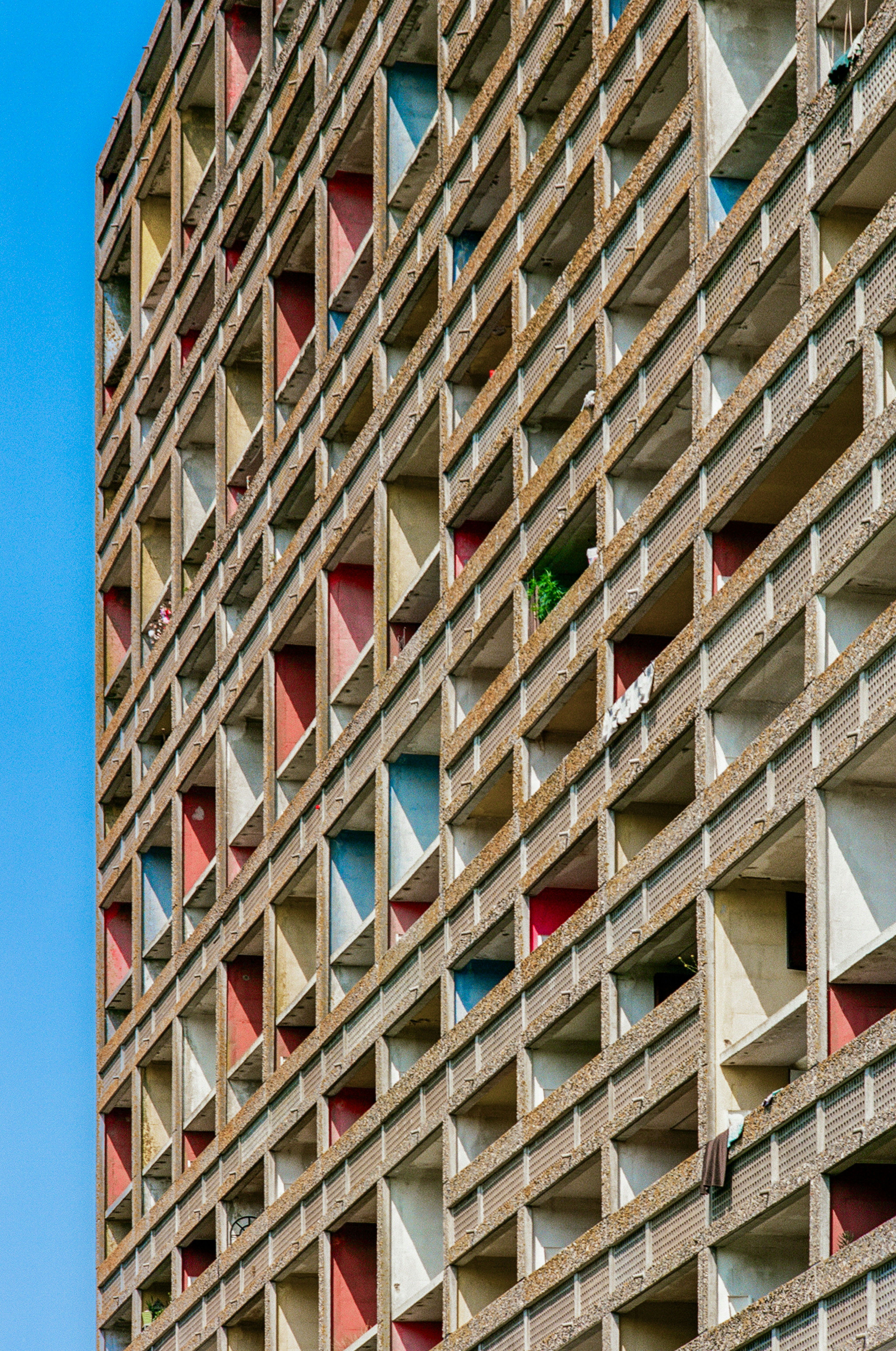 Photograph of the iconic Maison Radieuse in Rezé (near Nantes, France), designed by modernist architect Le Corbusier in the 1950s. The image captures the rhythmic geometry of the balconies and loggias in red, blue, and yellow tones, emblematic of brutalist architecture. Shot in July 2025 using a Nikon F3 HP with a Nikkor 105mm f/2.5 lens, on Kodak Portra 160 color film. | A concrete building shows numerous open windows.