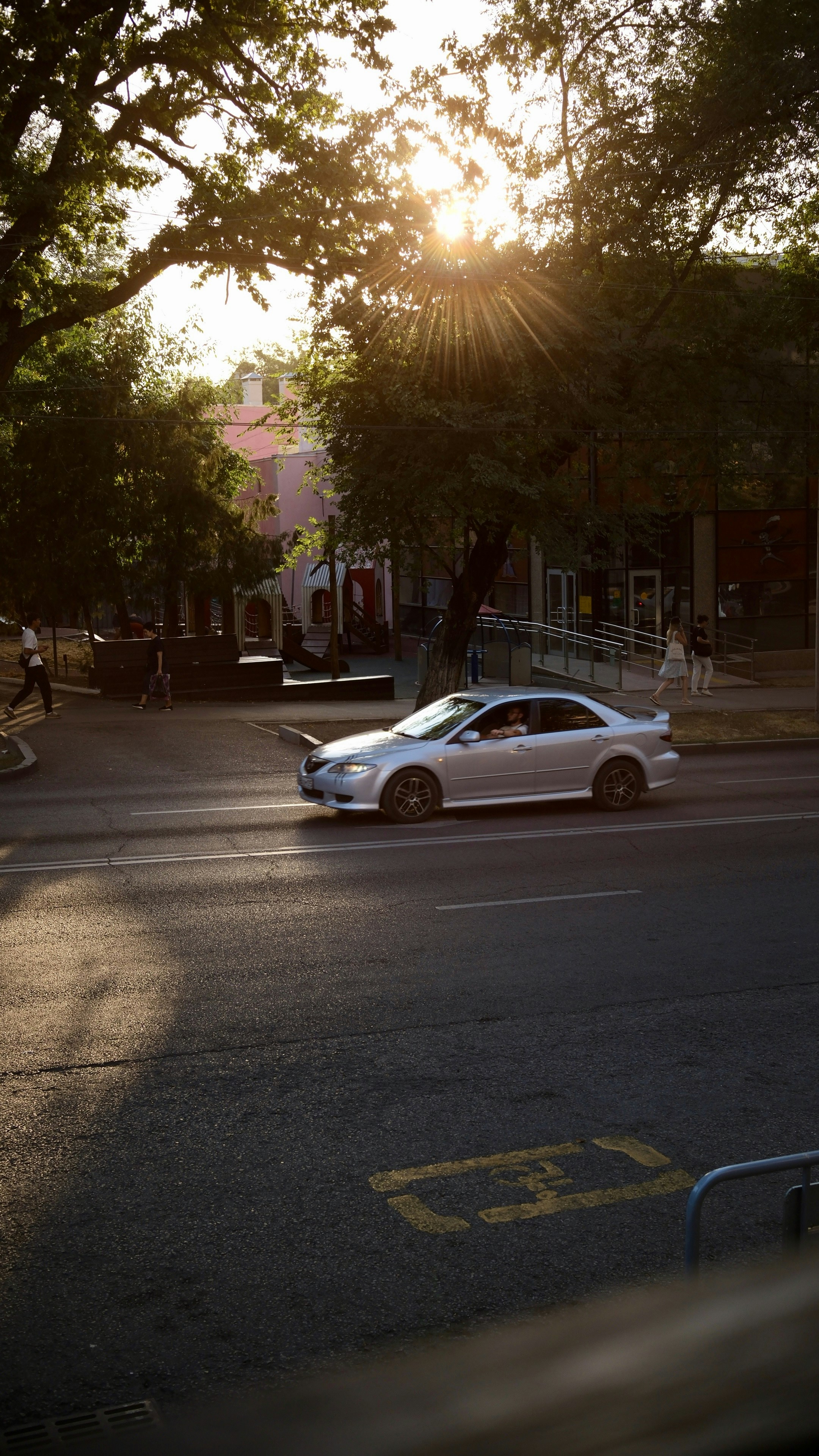 Silver car cruising down a sunlit street, framed by lush trees and a vibrant urban backdrop. The sun casts a warm glow, enhancing the evening atmosphere.