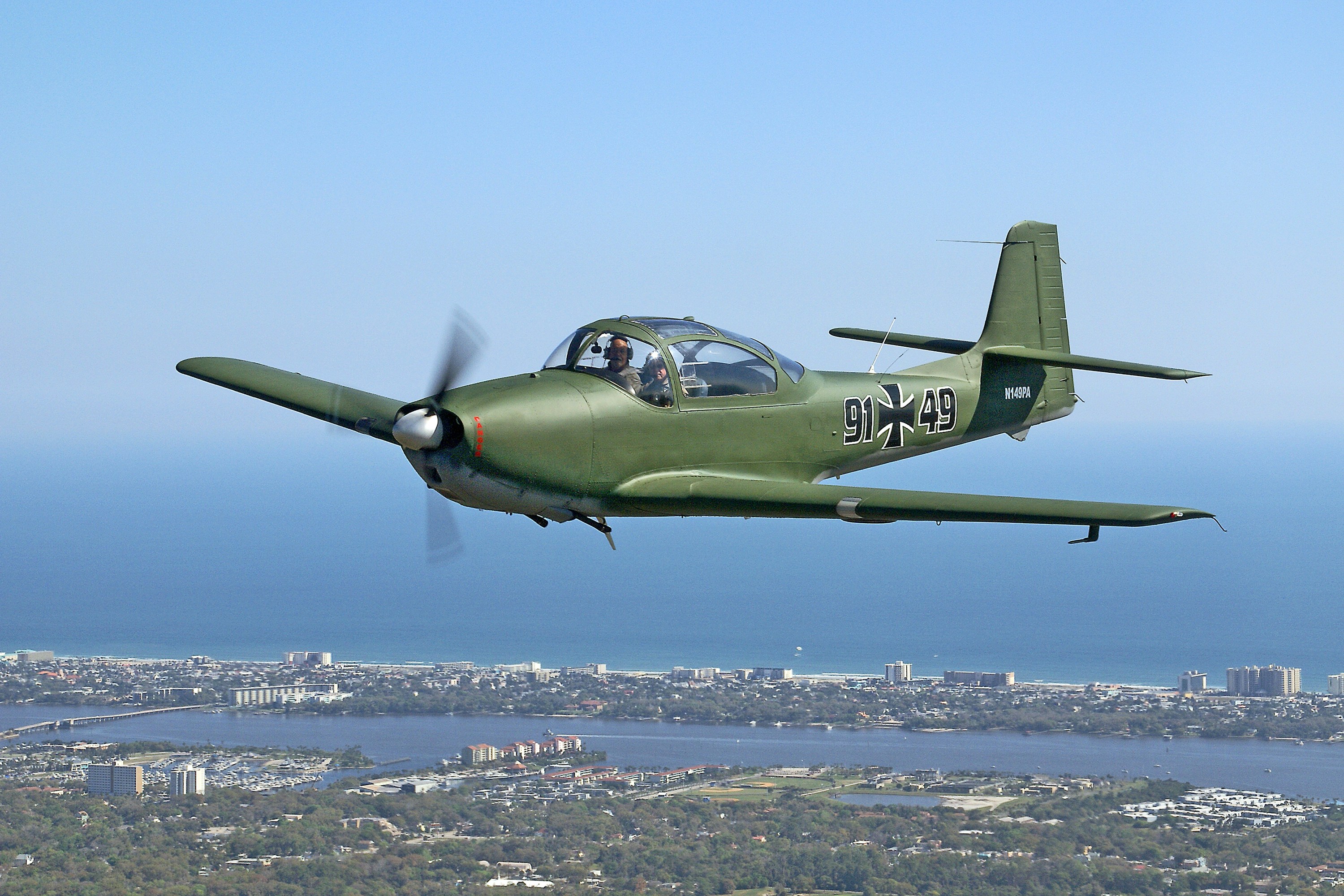 A Fouck-Wulf P-149 flying over the East Coast of Florida | An olive green airplane flies above the city.