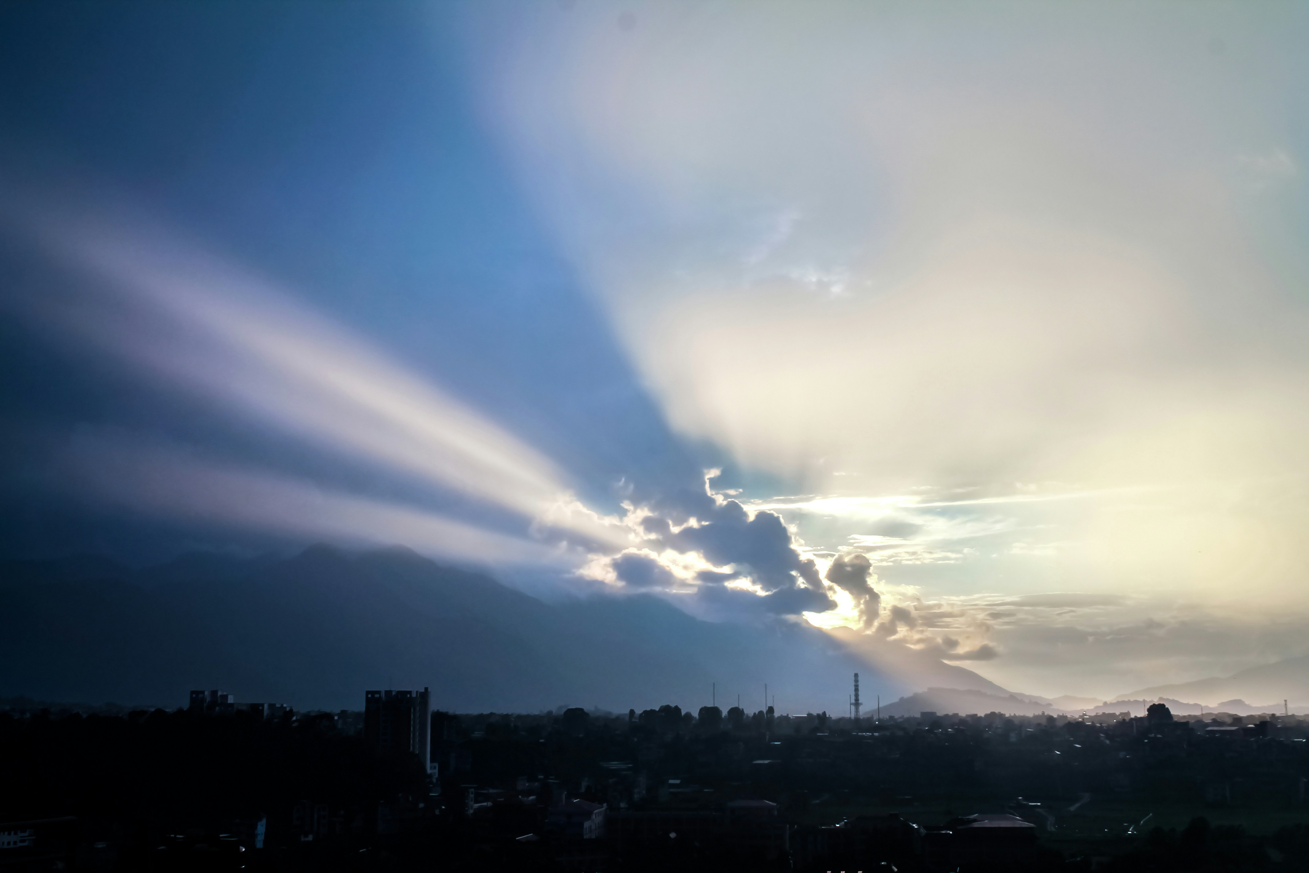 Golden Rays Through Grey Skies: Nepal’s Cloudy Sun | Sunbeams shine through the clouds over a city.