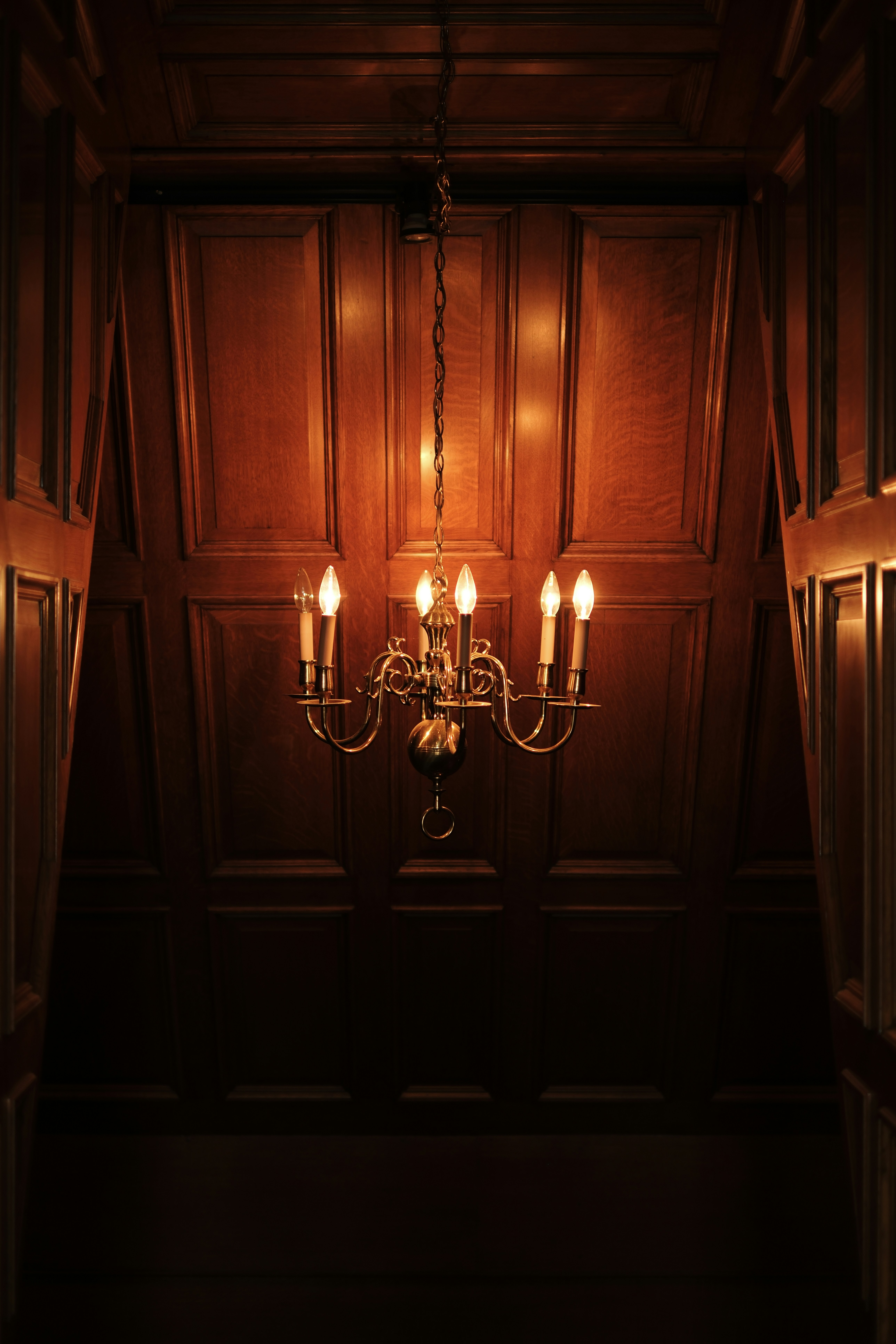 A vintage chandelier hangs gracefully from a wooden ceiling, casting a warm glow in an intimate space. The intricate design contrasts beautifully with the rich wood paneling.