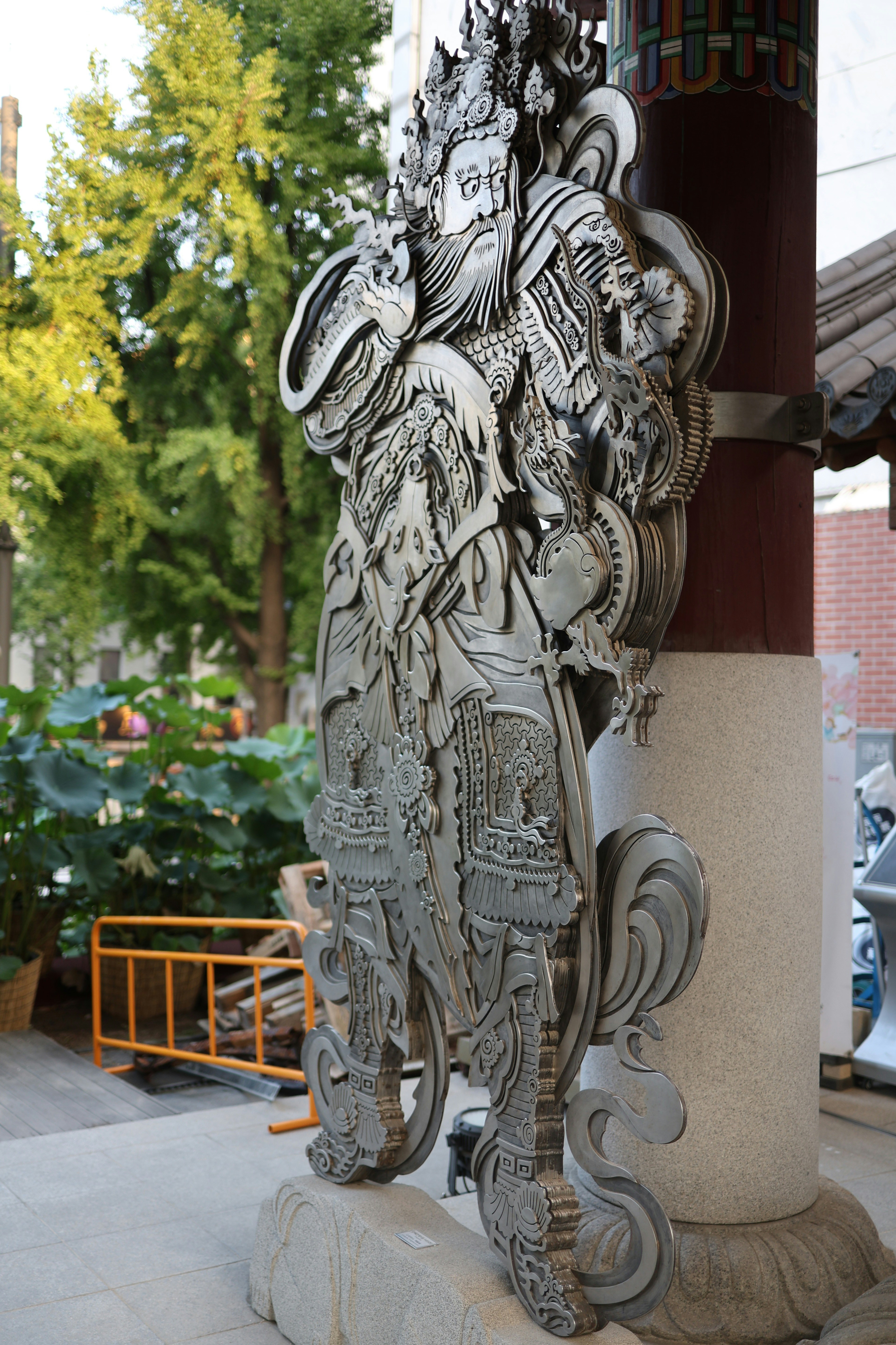 Intricate silver sculpture of a guardian deity standing beside a pillar, surrounded by lush greenery. The detailed craftsmanship highlights cultural significance.