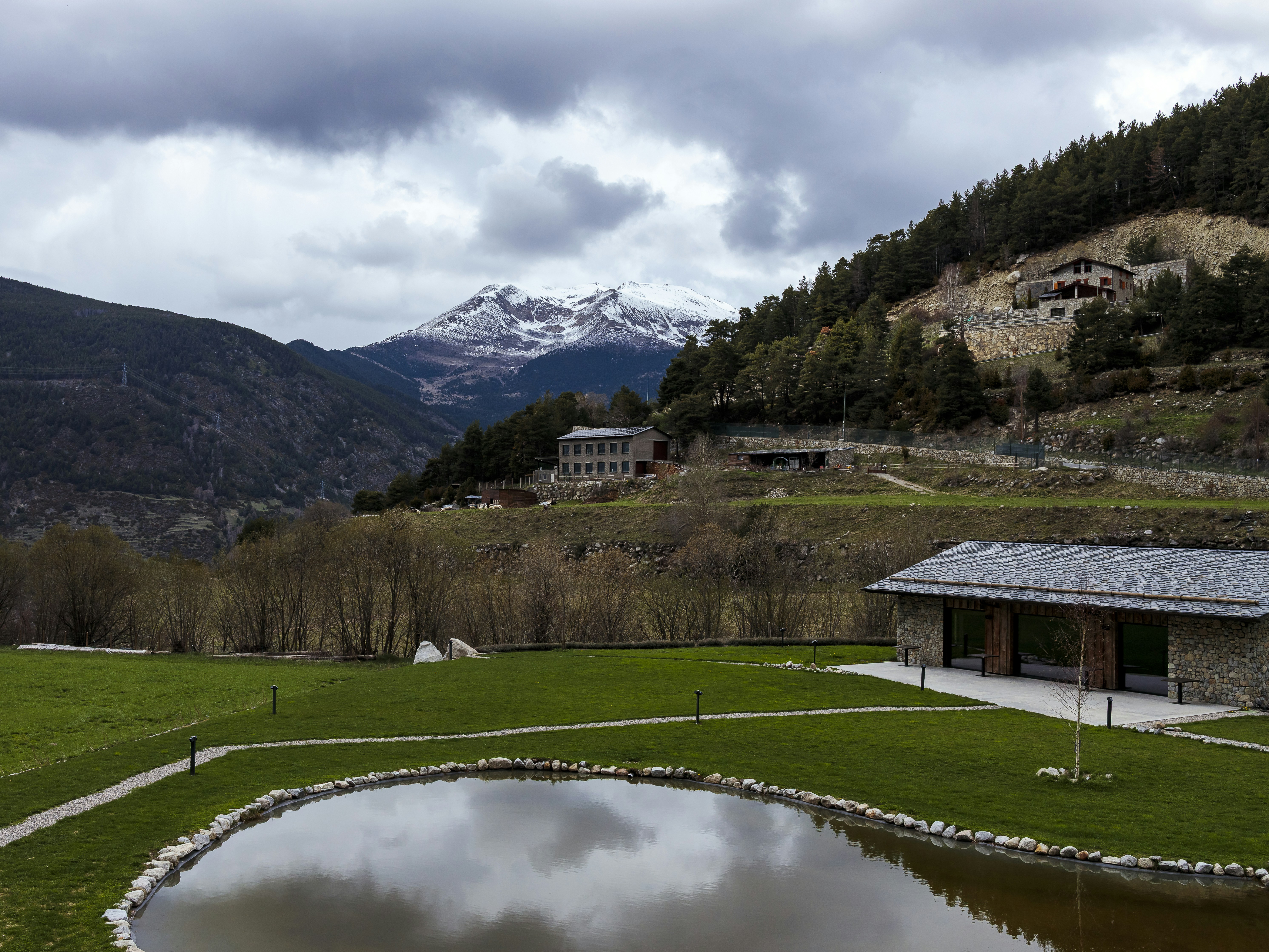 A peaceful mountain landscape featuring a small pond reflecting the cloudy sky, modern building nearby, rolling green fields, and snow-capped peaks in the background. The scene exudes tranquility and natural beauty, typical of a mountain region during early spring or late winter. | Mountain landscape featuring a building and a pond.