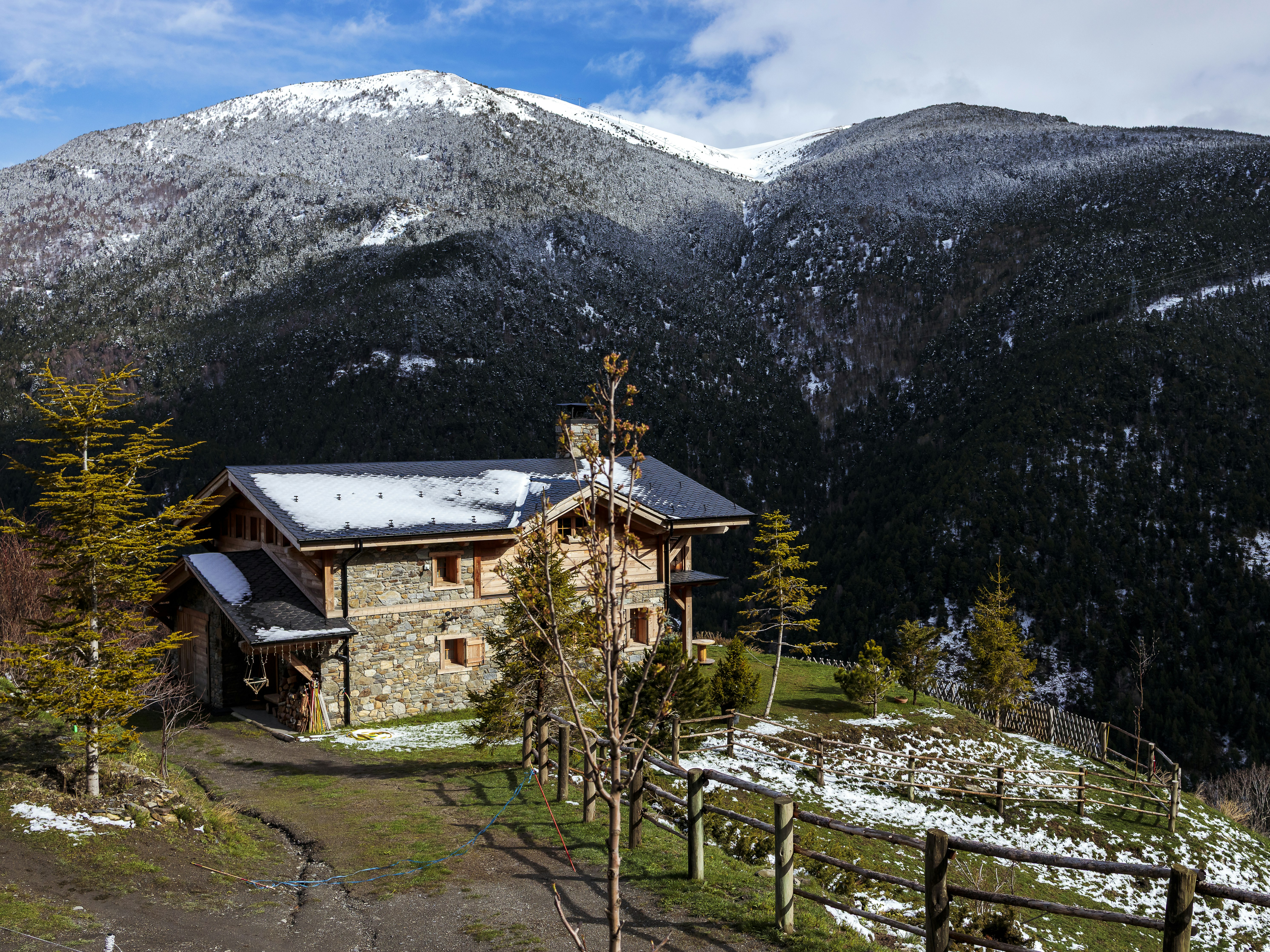 A picturesque mountain scene showcasing a charming stone and wood cabin situated on a lush, partially snow-covered hillside. Tall pine trees surround the cabin, leading to majestic, snow-capped mountains in the background. The scene radiates tranquility and the beauty of remote mountain living during late winter or early spring, under a partly cloudy sky. | A snowy mountain home resides among nature.