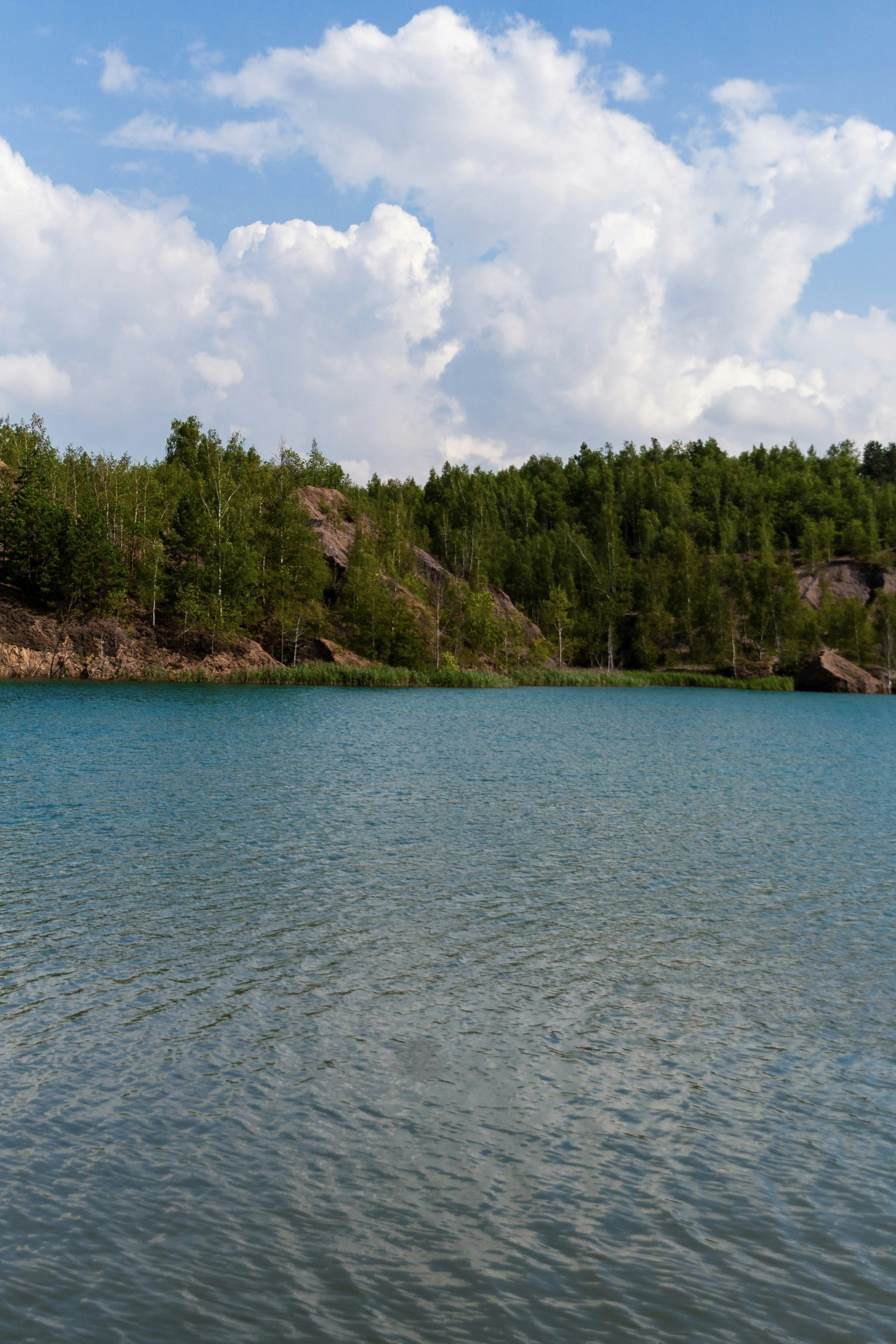 A vertical shot of the calm turquoise water of the lake with the opposite shore featuring a line of trees on a brown hill under a partly cloudy, blue sky | Turquoise lake with trees and a cloudy sky.
