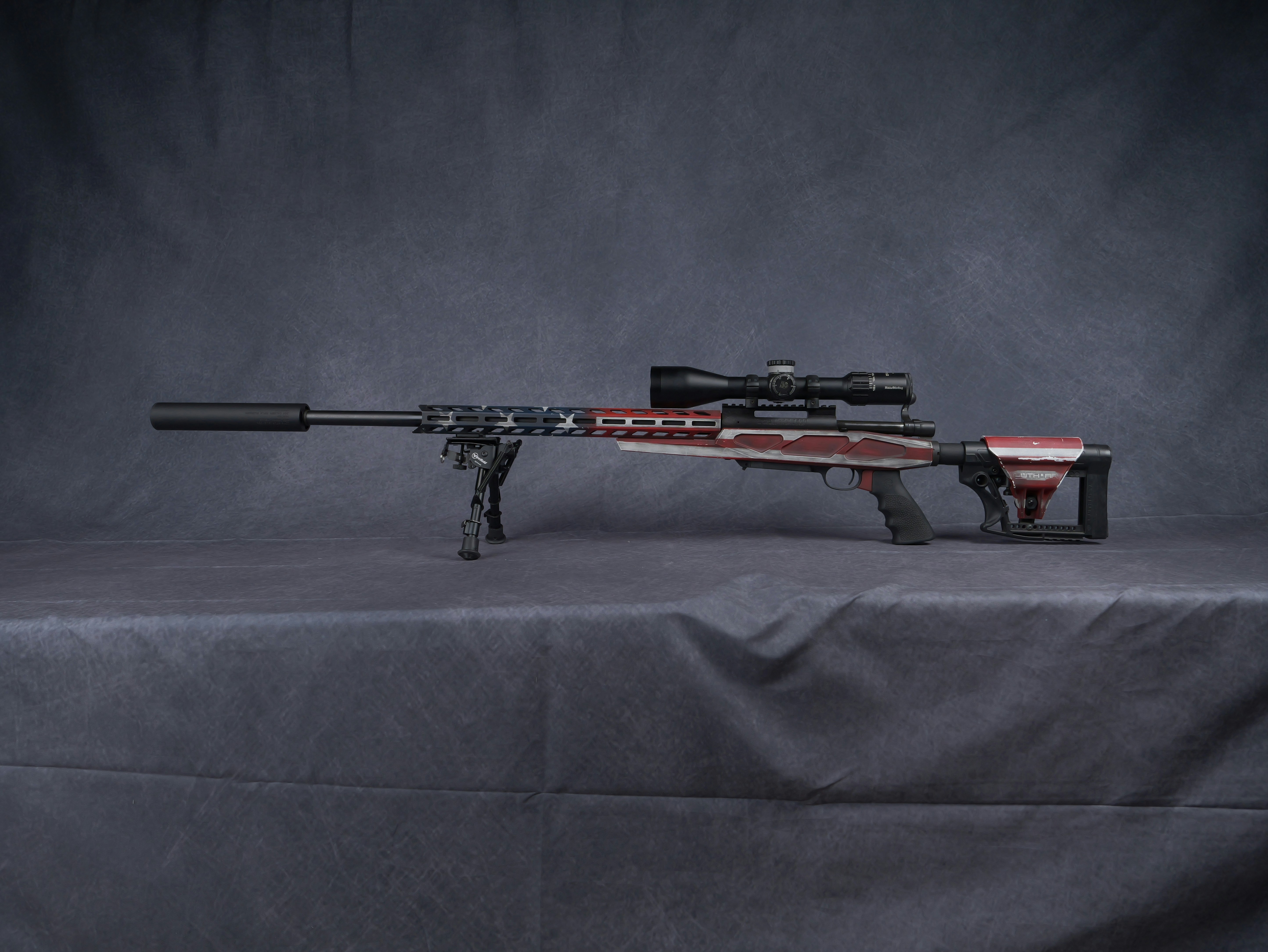 A sleek, modern rifle with a suppressor and advanced scope, set against a textured gray backdrop.
