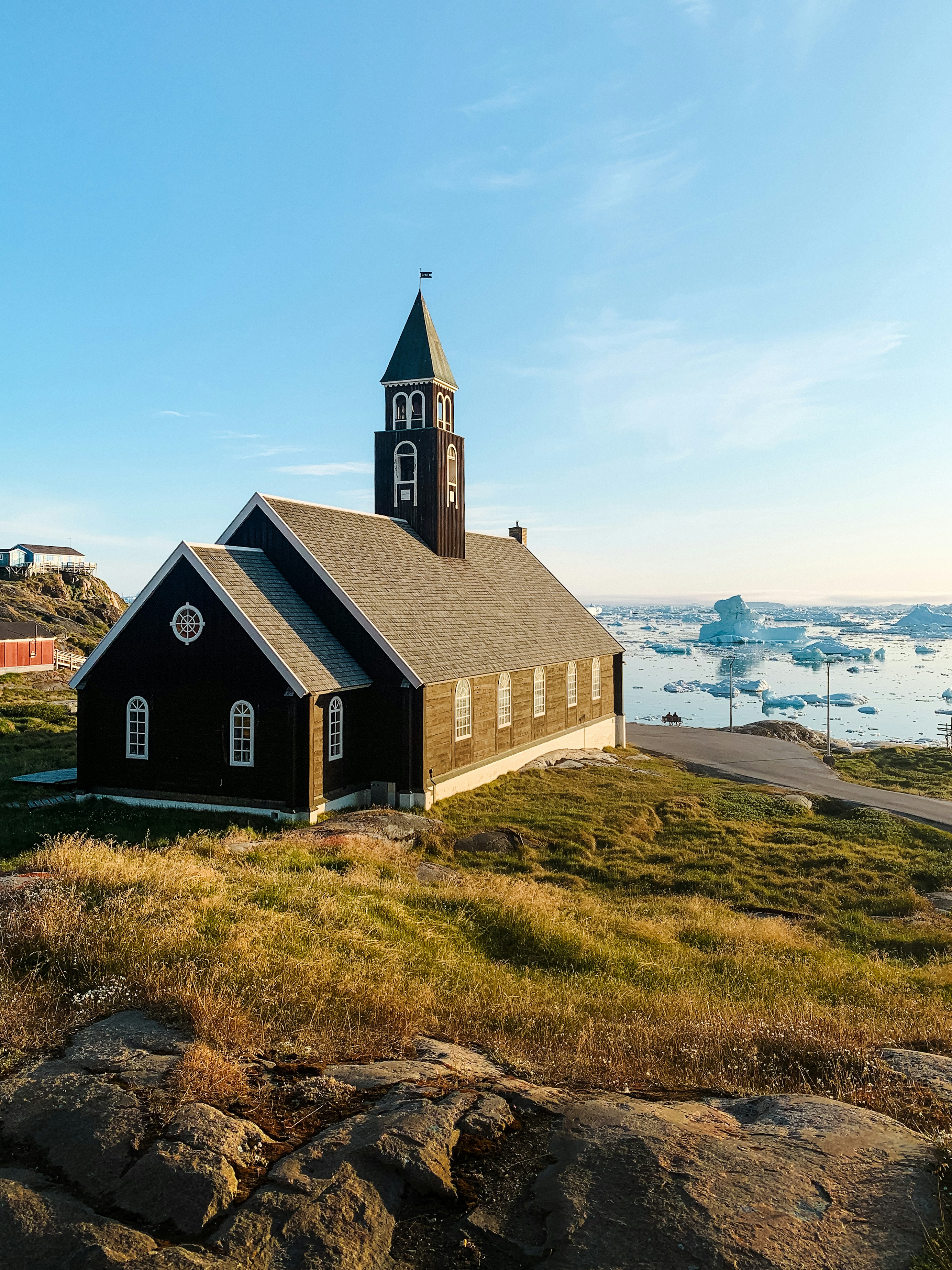 The iconic black church of Ilulissat, Greenland, a beacon against the arctic landscape. This historic chapel stands by the UNESCO Icefjord, a testament to resilient faith and Nordic heritage. | A black church overlooks a sea of icebergs.