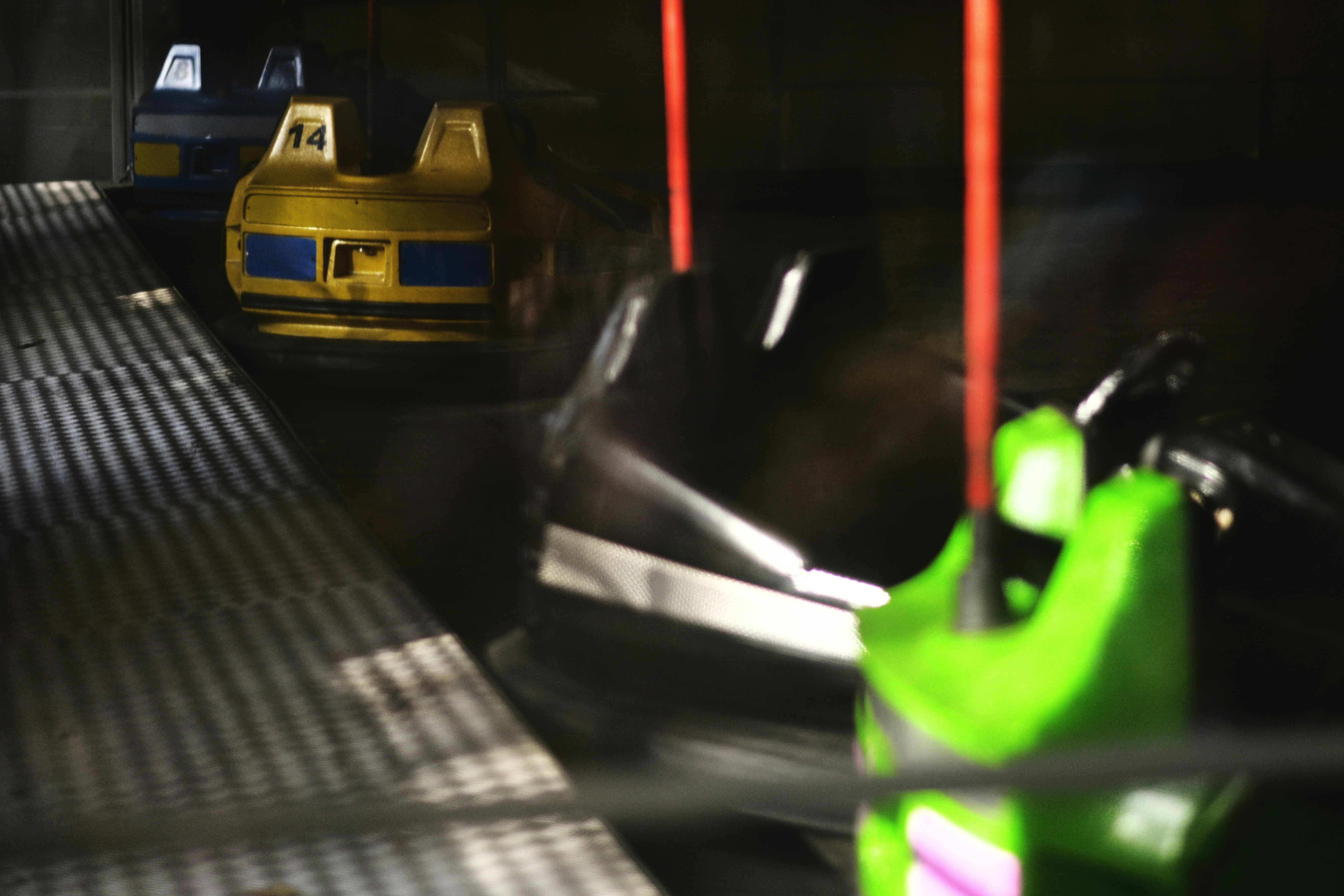 A vibrant green bumper car, captured in motion, dominates the foreground. Its bright color contrasts with the darker, slightly blurred background of the bumper car arena. A yellow bumper car, marked with the number 14, is parked in the distance, while a second, blue car is visible behind it. The red support poles of the ride and the textured metal floor add to the dynamic, energetic feel of the scene, evoking the chaotic fun of an amusement park. | Bumper cars sit idle in a darkened space.