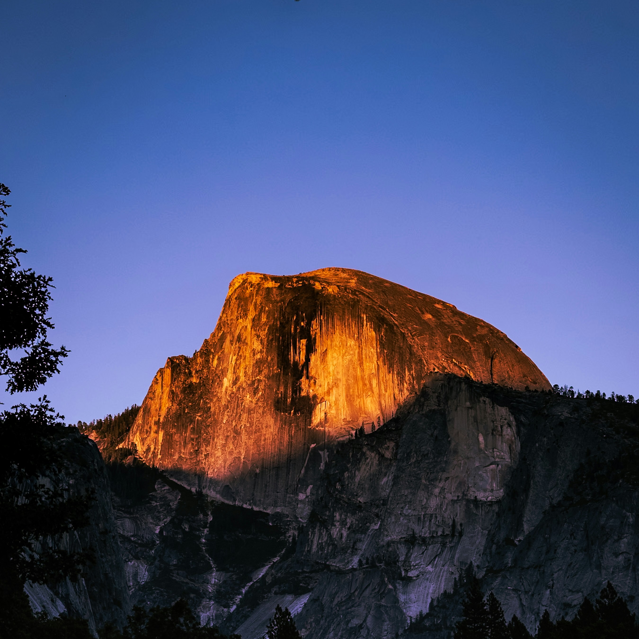 Half Dome illuminated by the warm light of sunset, surrounded by a twilight sky and rugged mountain terrain.