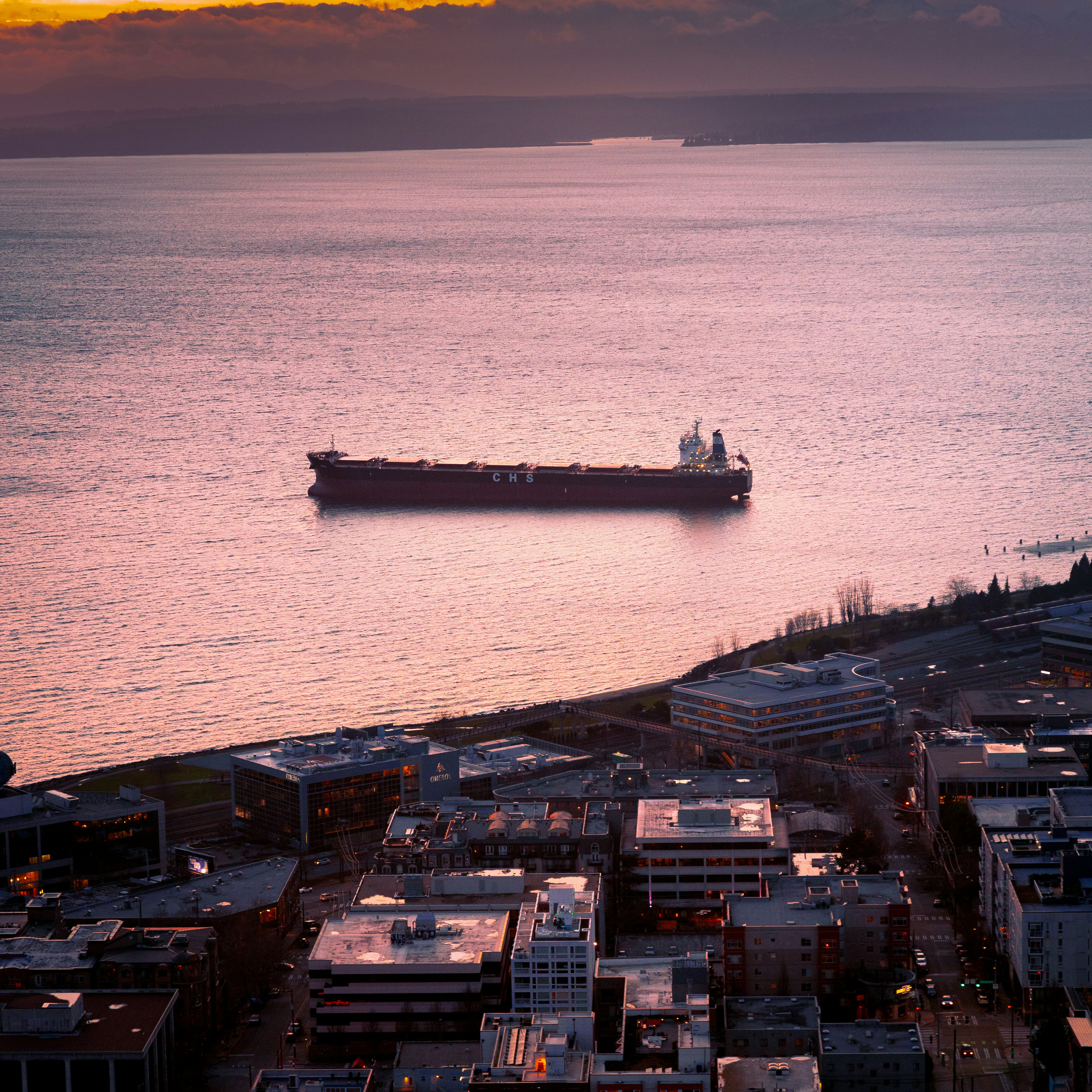 Cargo ship navigating tranquil waters at sunset, with urban landscape in the foreground. Soft hues of twilight reflect on the water's surface.